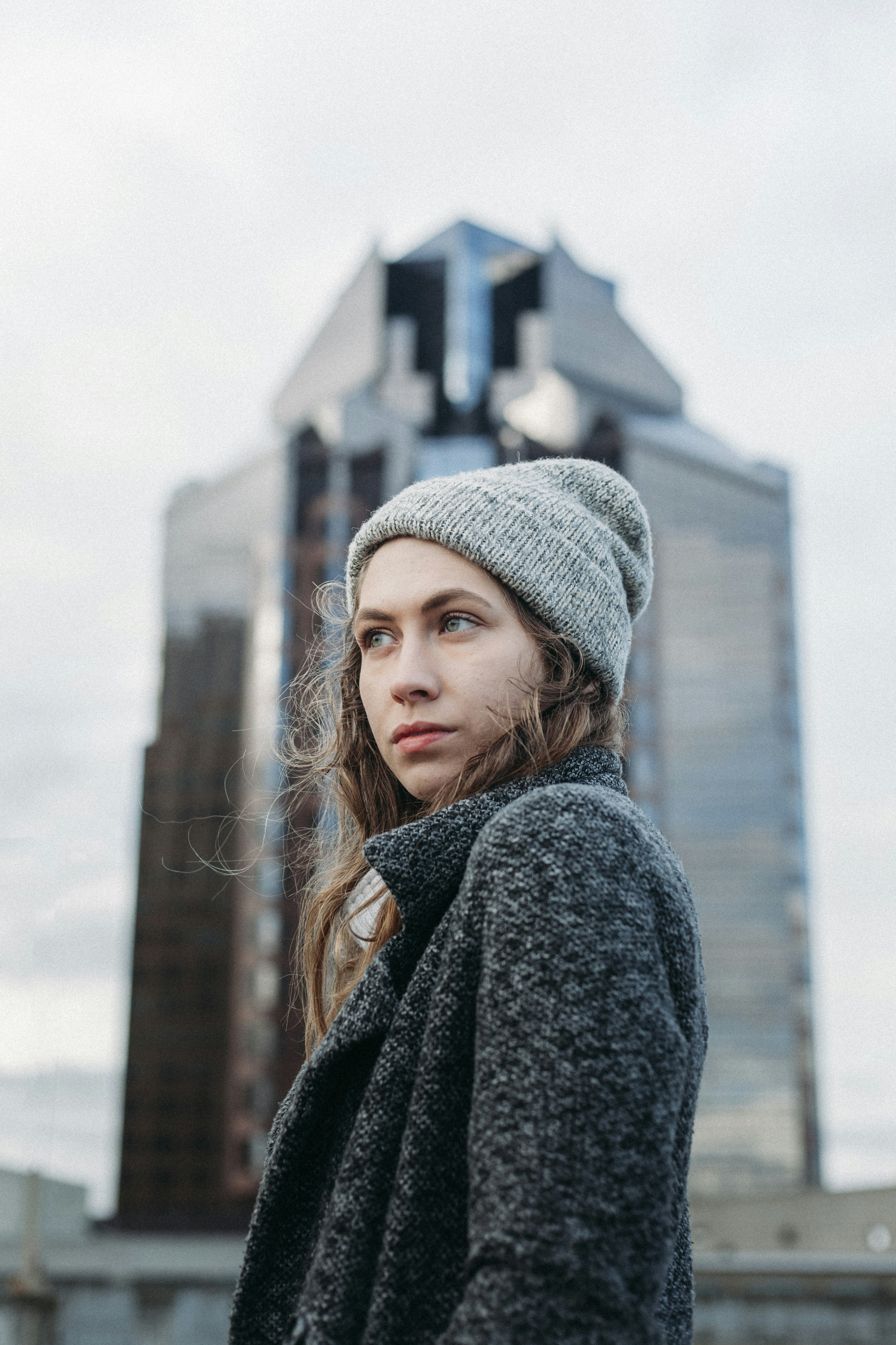 Young woman in a gray beanie gazes thoughtfully against a backdrop of modern architecture. Wind tousles her hair, adding to the contemplative mood.