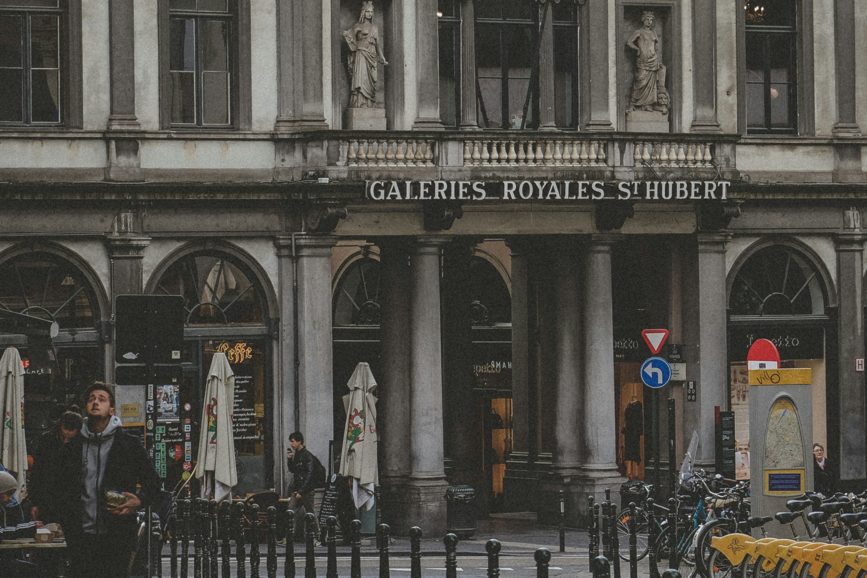 Historic shopping arcade featuring ornate architecture and bustling activity, with outdoor seating and pedestrians. The grandeur of the Galeries Royales St Hubert is on full display.