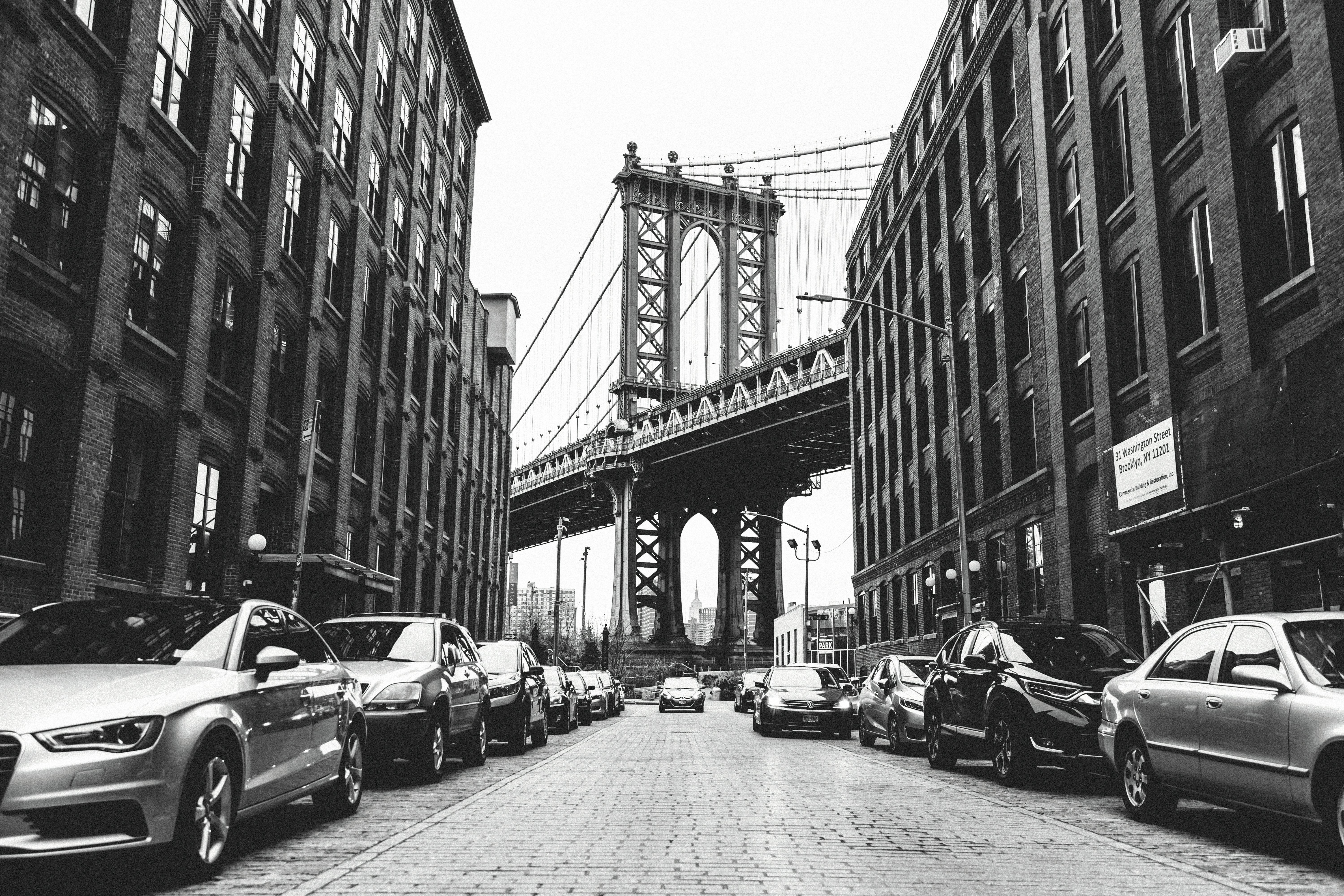 Grayscale view of a city street with parked cars and a bridge framed by tall buildings.