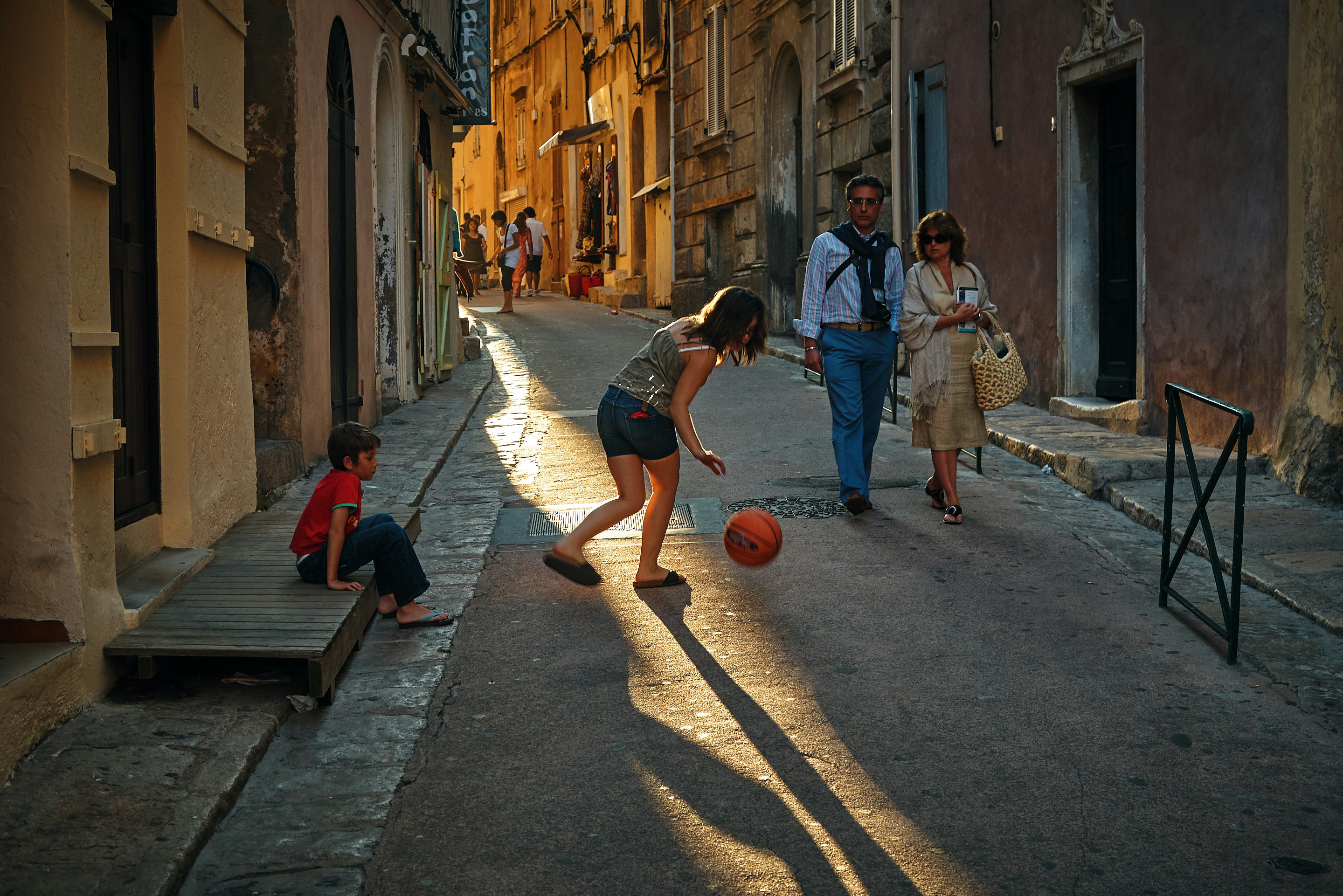 A girl dribbles a basketball on a sunlit cobblestone street while a boy sits nearby, watching. Two adults stroll past, adding life to the charming alleyway.