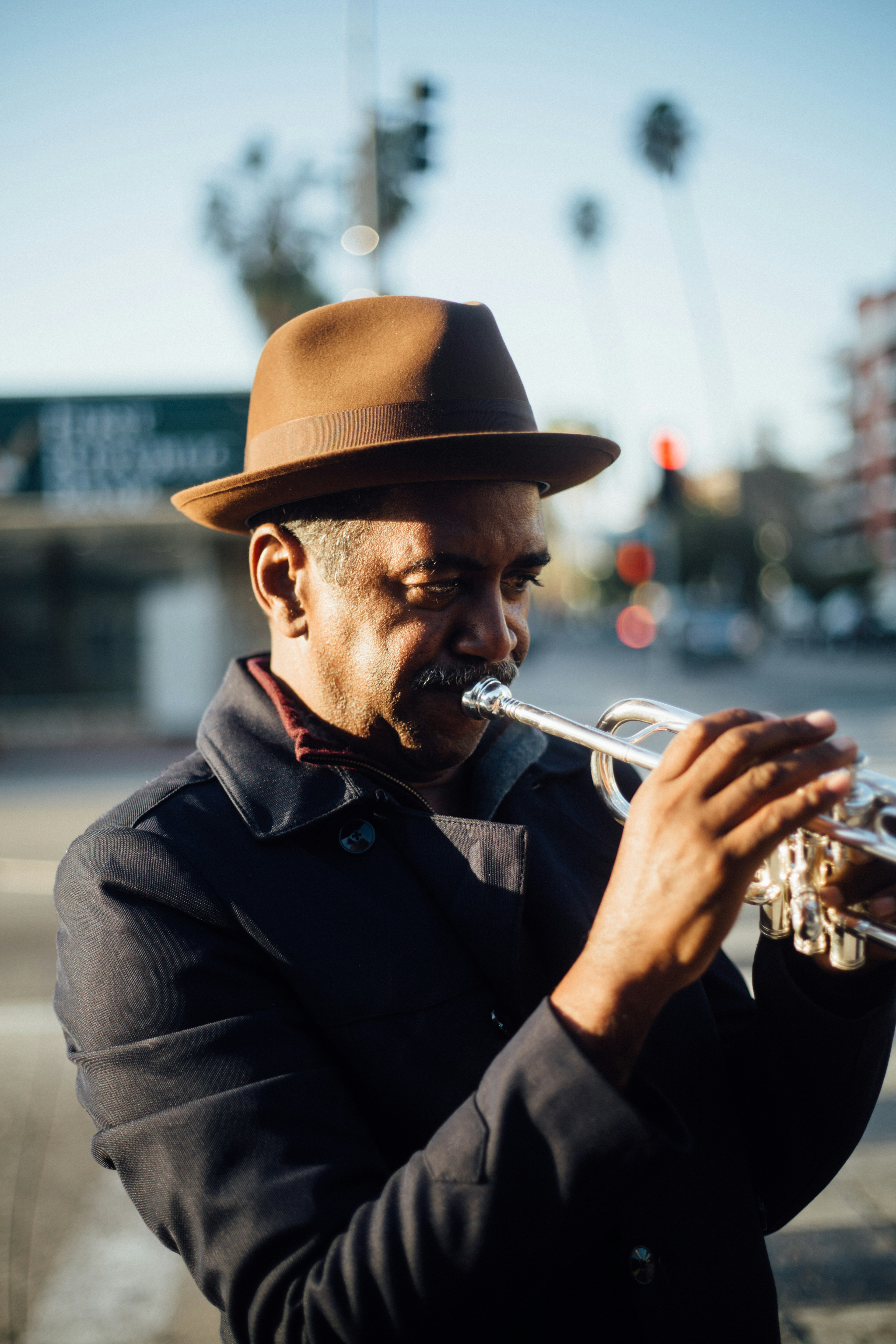 Musician passionately playing the trumpet on a bustling street, with palm trees and traffic lights in the background.