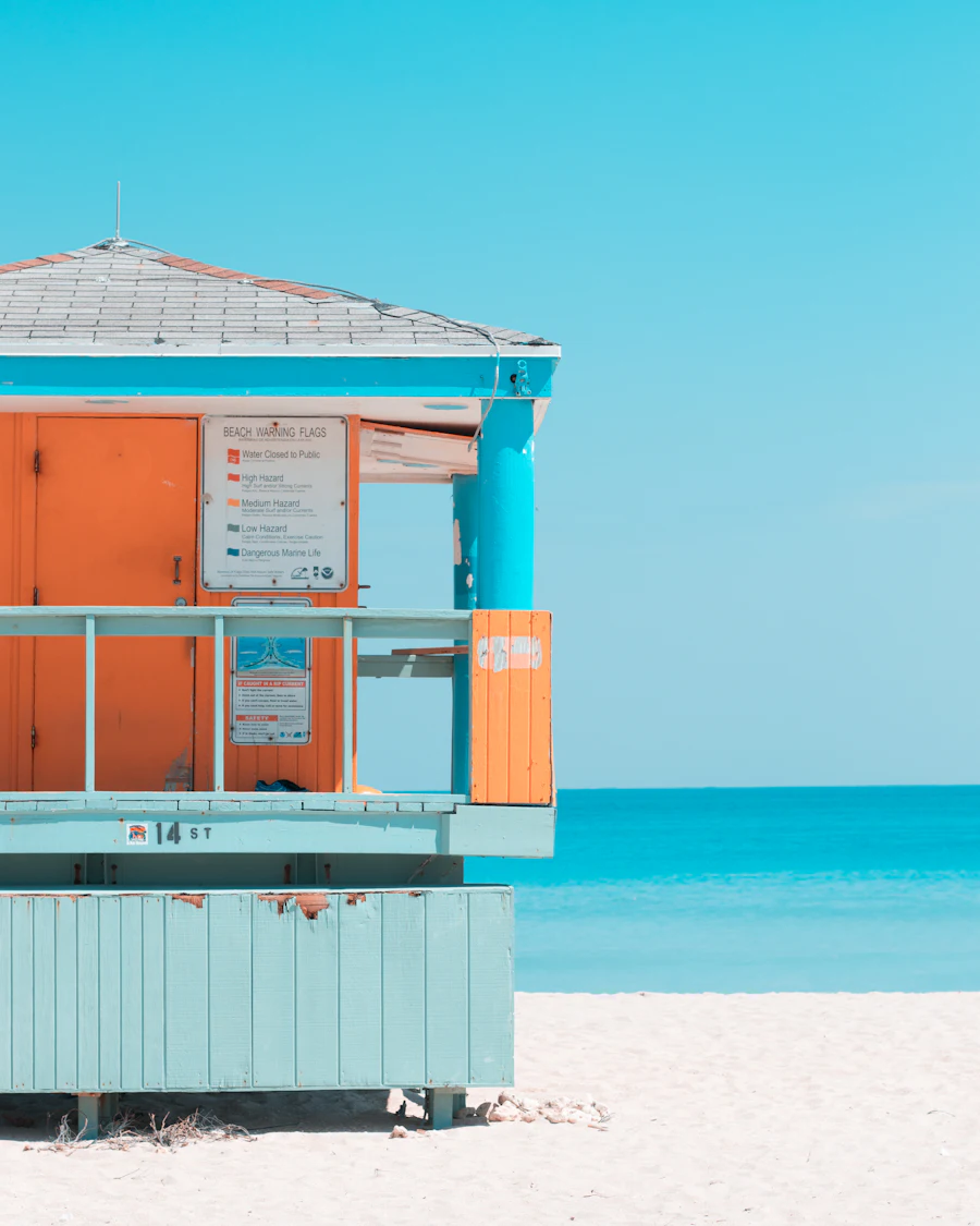 Turquoise and orange lifeguard hut beside a bright white beach with calm blue ocean and sky. The structure sits left of frame with open water stretching across the right side.