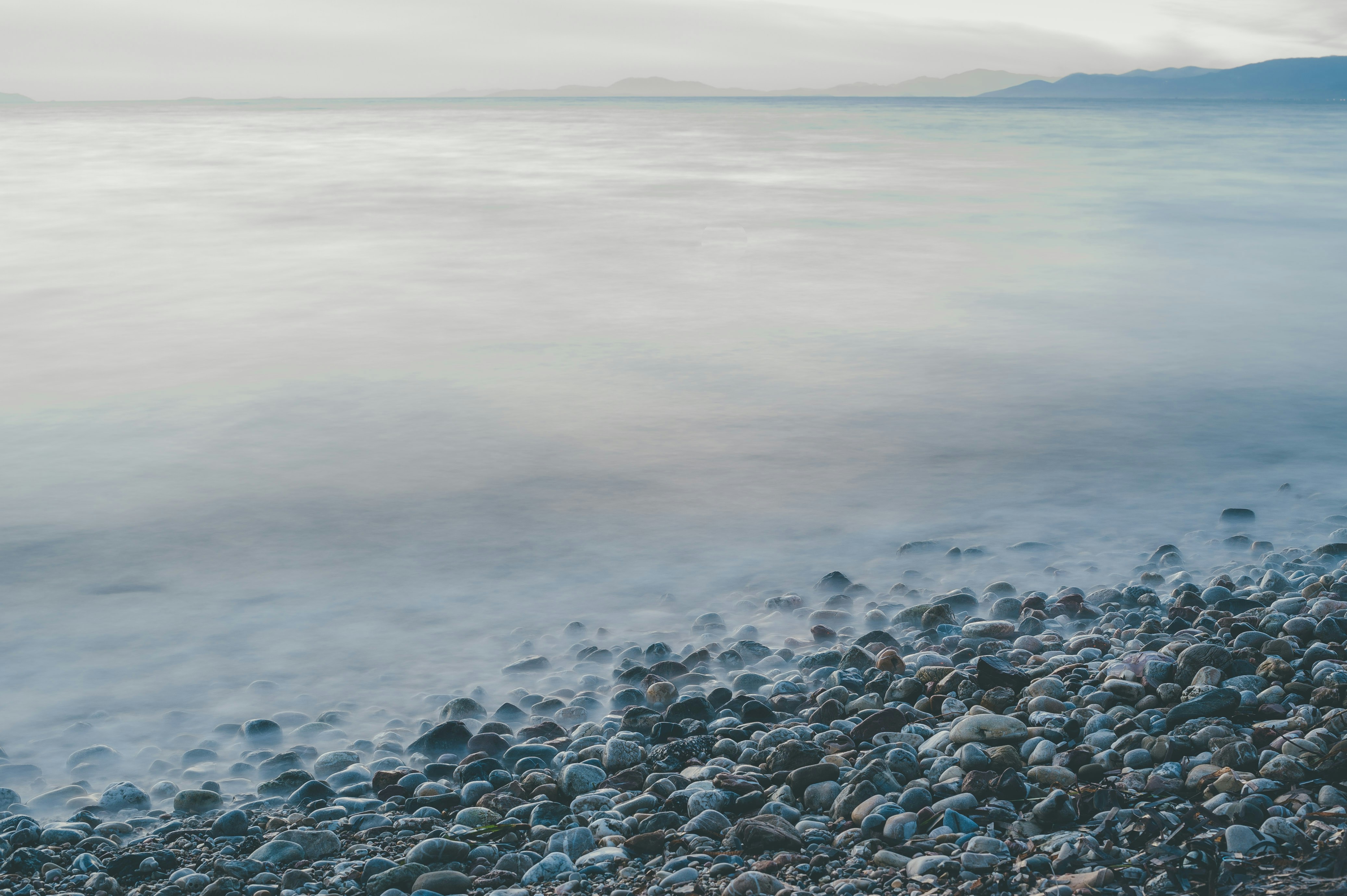 Smooth pebbles meet the gentle, misty waves of a tranquil shoreline under a soft, overcast sky.