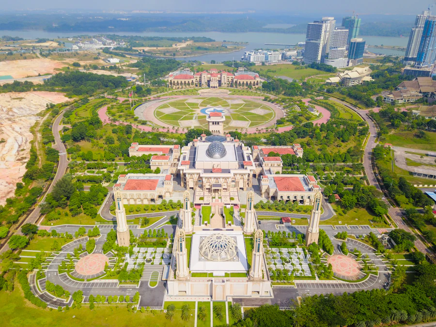 Symmetrical palace-like complex with a large circular plaza, domed central building, and landscaped roads surrounded by trees. High-rise towers and a river sit in the distance under a hazy sky.