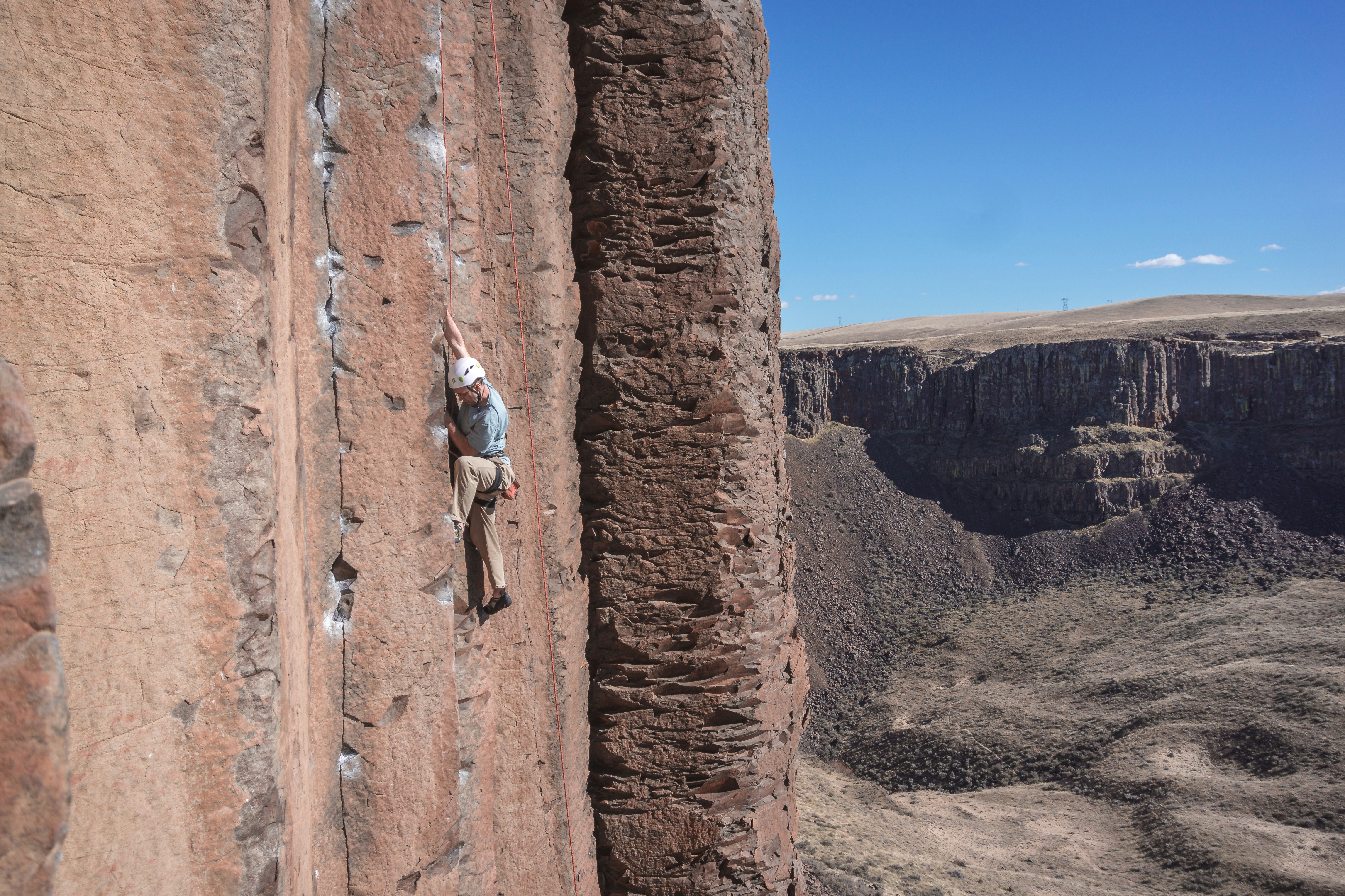 Climber scaling a vertical rock face against a vast canyon backdrop under a clear blue sky.