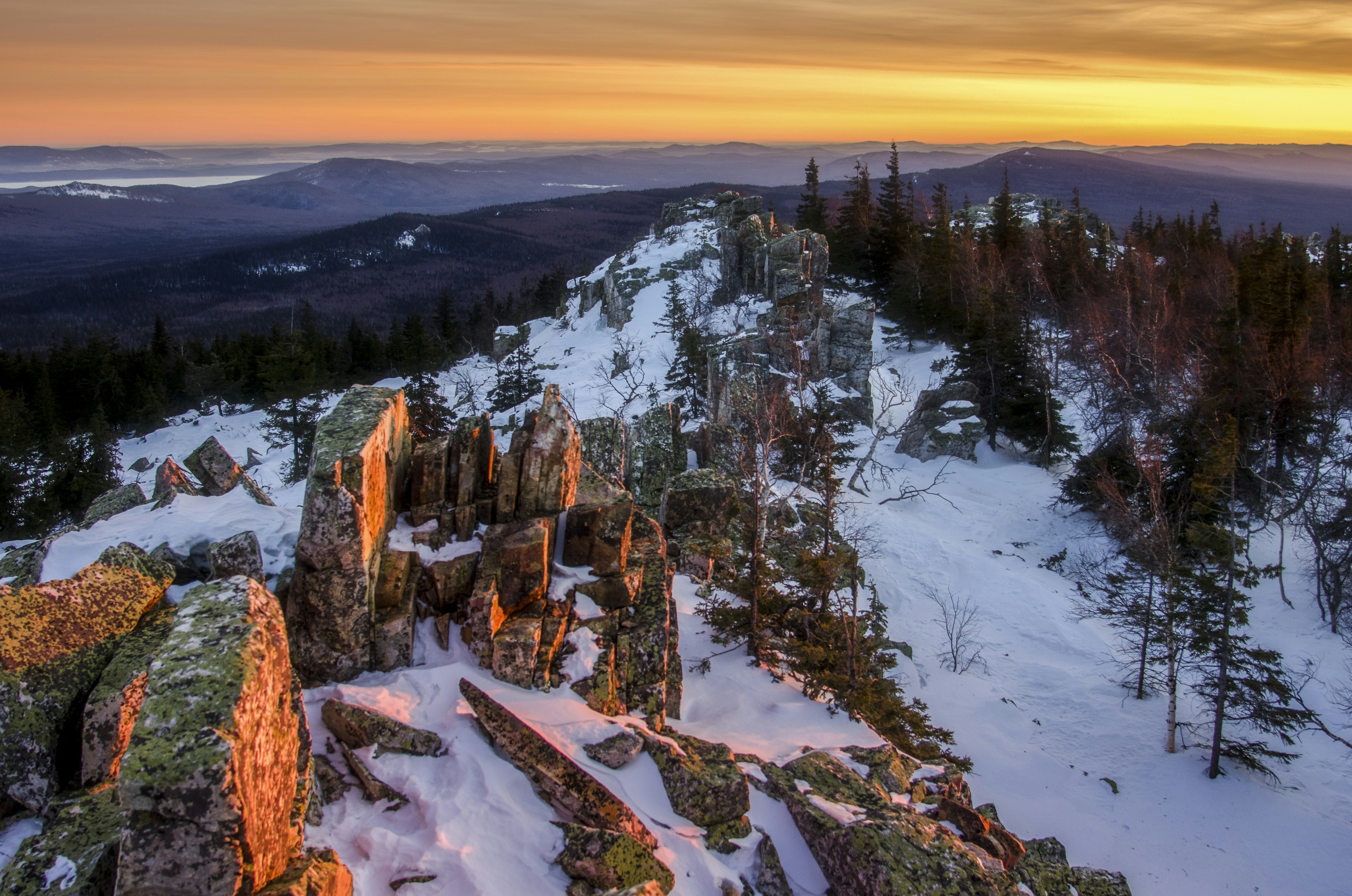 Snow-covered rocky outcrop overlooking a vast mountainous landscape during sunset, with soft hues illuminating the scene.