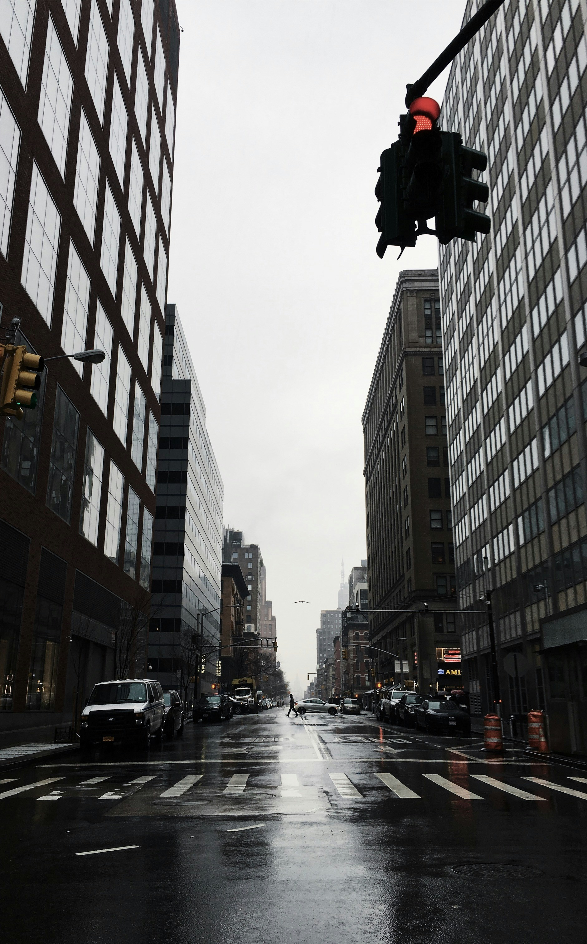 Rain-soaked urban street flanked by glass towers, with a red traffic light overhead and a wet, reflective road receding toward a distant vanishing point.