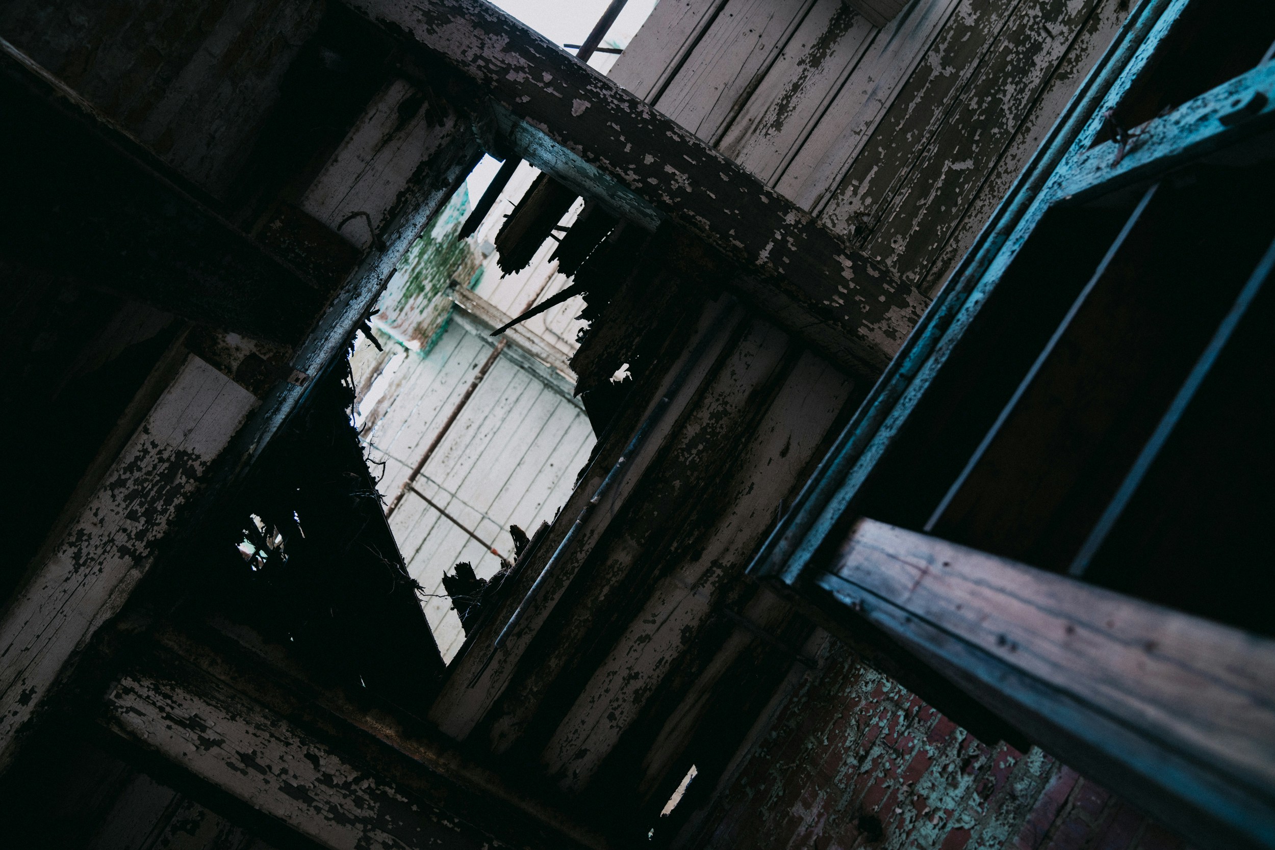 View from below an abandoned structure, revealing a gaping hole in the ceiling framed by weathered beams and peeling paint.