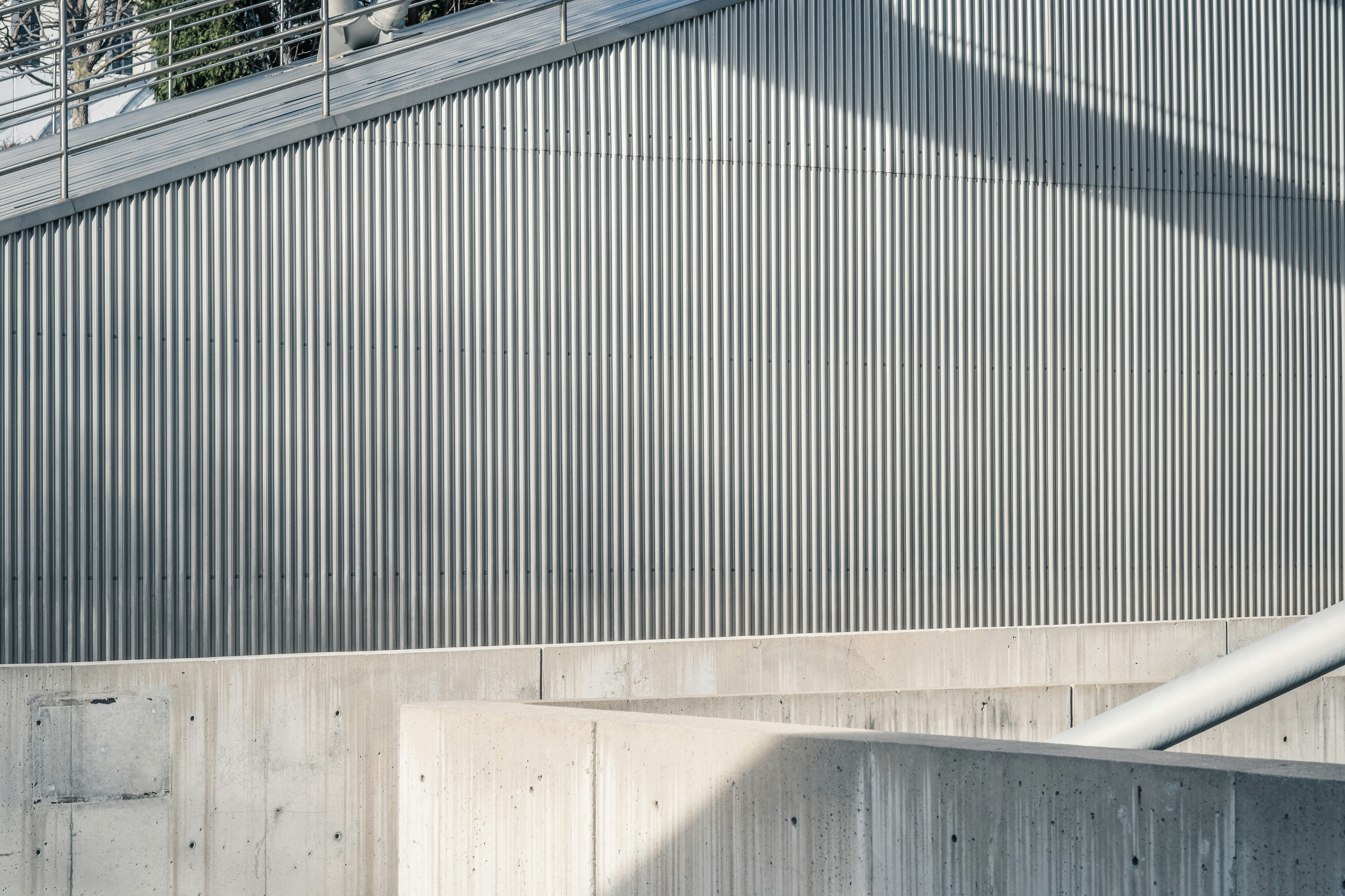 Gray corrugated metal facade with intersecting shadows and concrete elements.