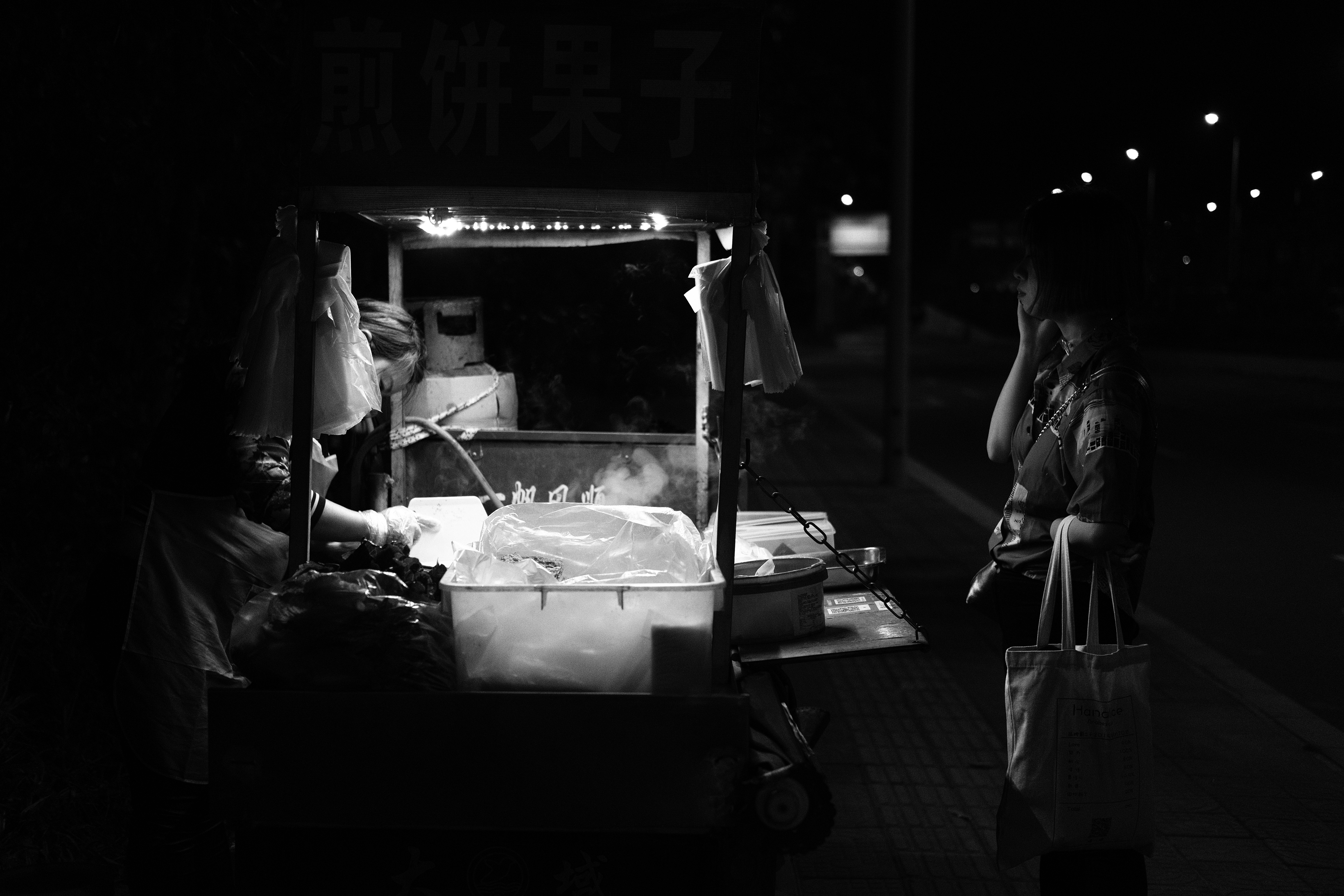 A street vendor prepares food at night, illuminated by a warm light, while a customer waits nearby. The scene captures the essence of urban nightlife.