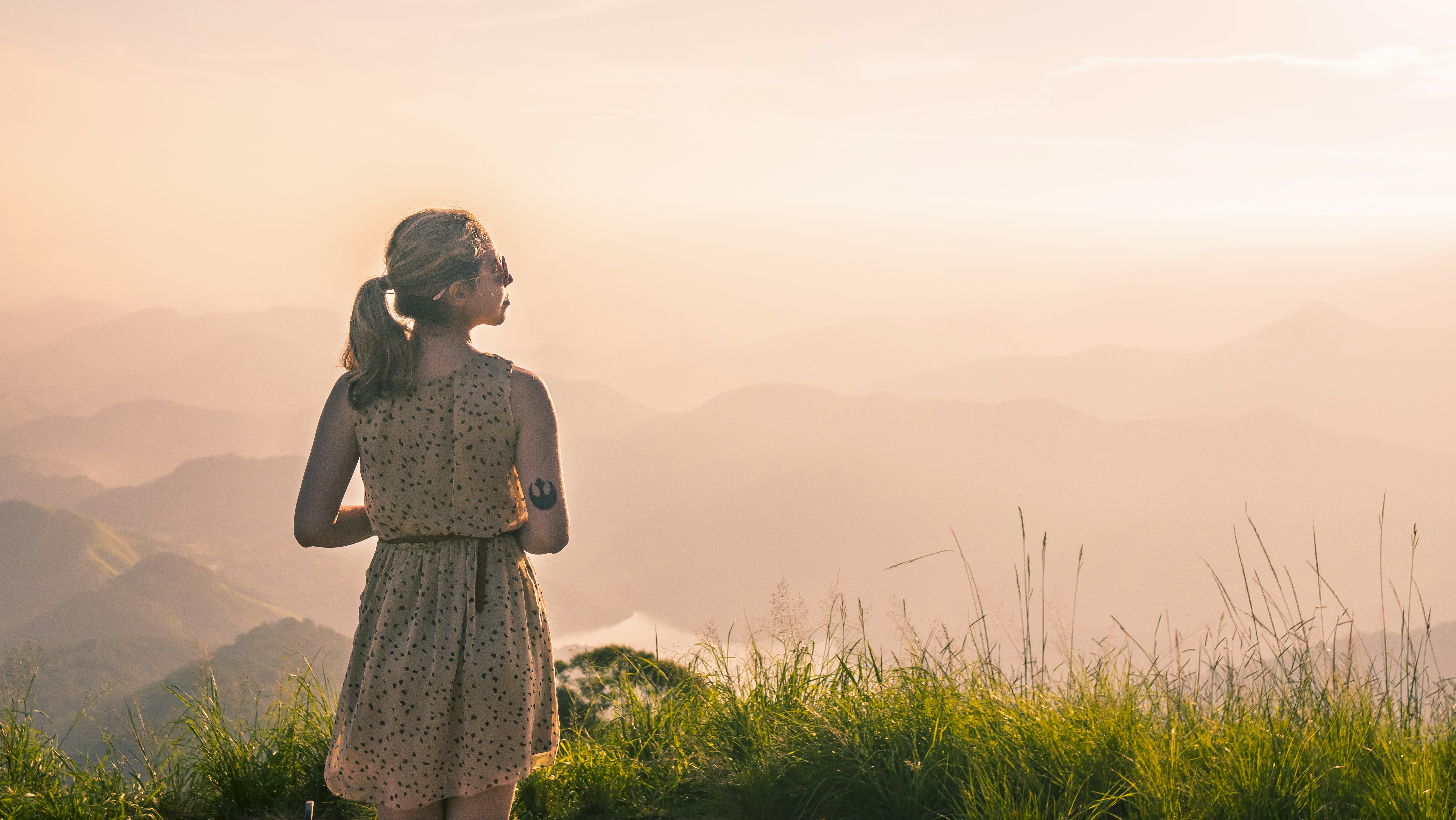 Woman standing in a grassy meadow gazing at a hazy sunrise over distant mountains.