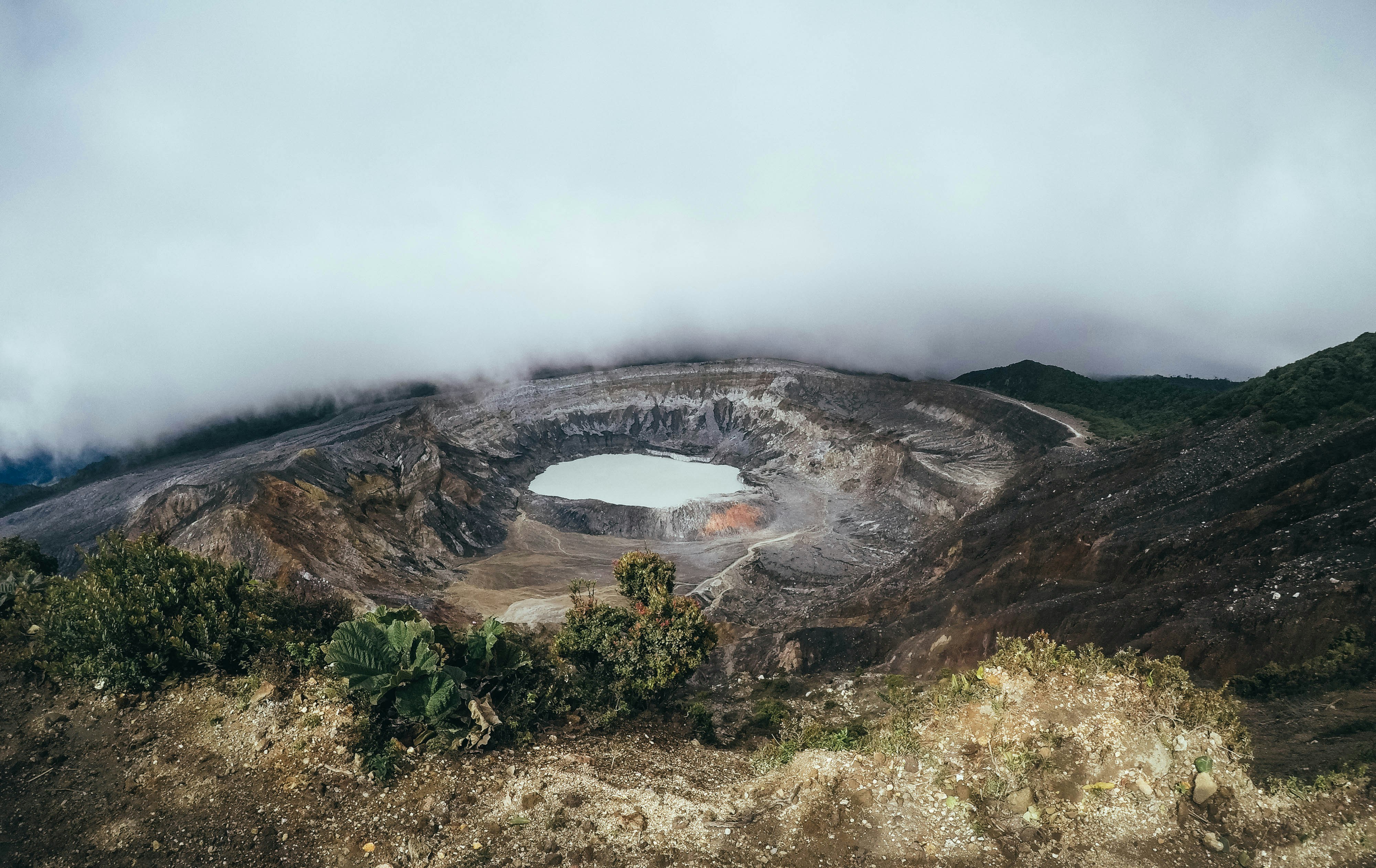 Volcanic crater with a sulfuric lake surrounded by rugged terrain and low clouds.