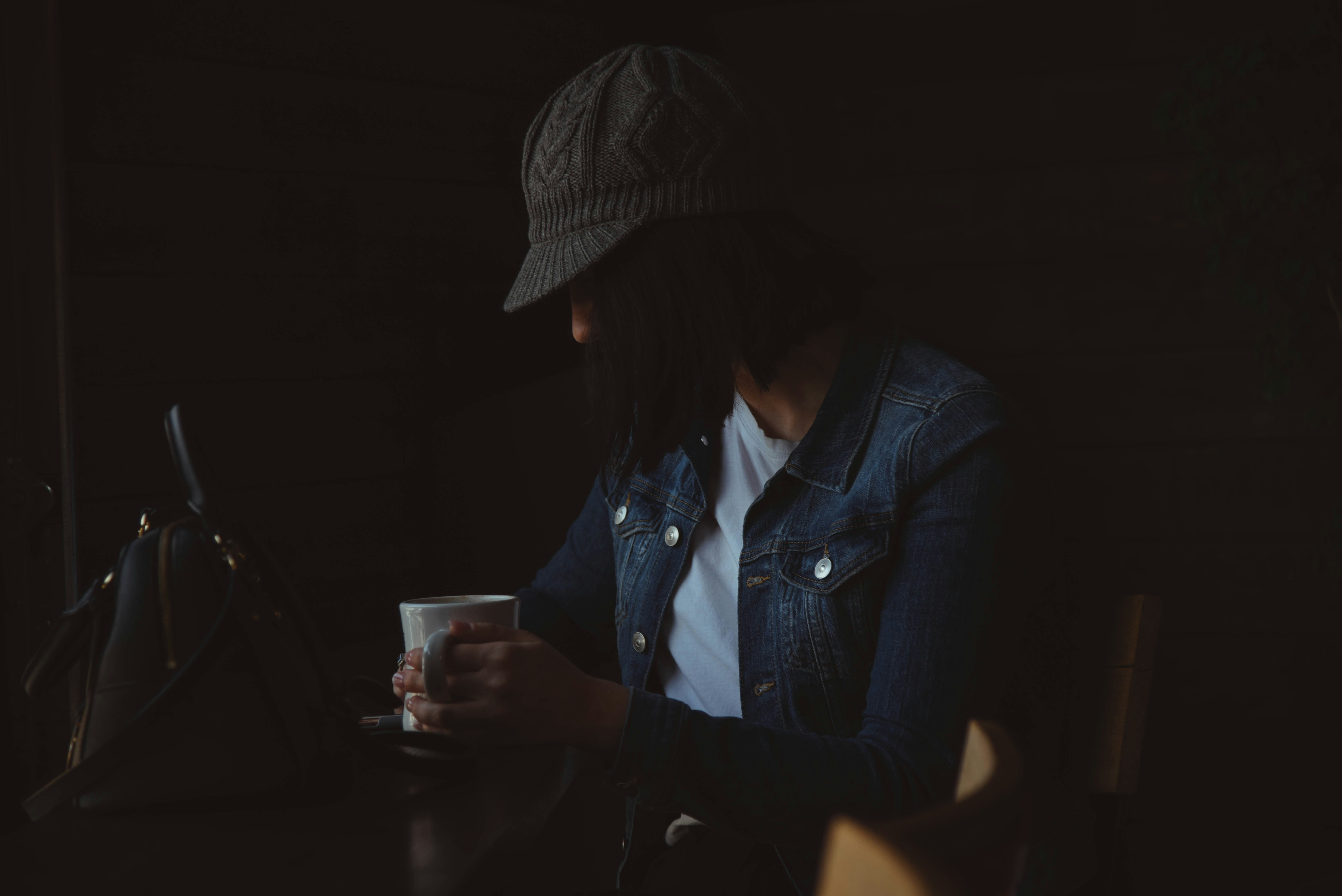Woman in a cozy setting, holding a cup, dressed in a denim jacket and hat, immersed in thought. The scene captures a serene atmosphere with soft lighting.