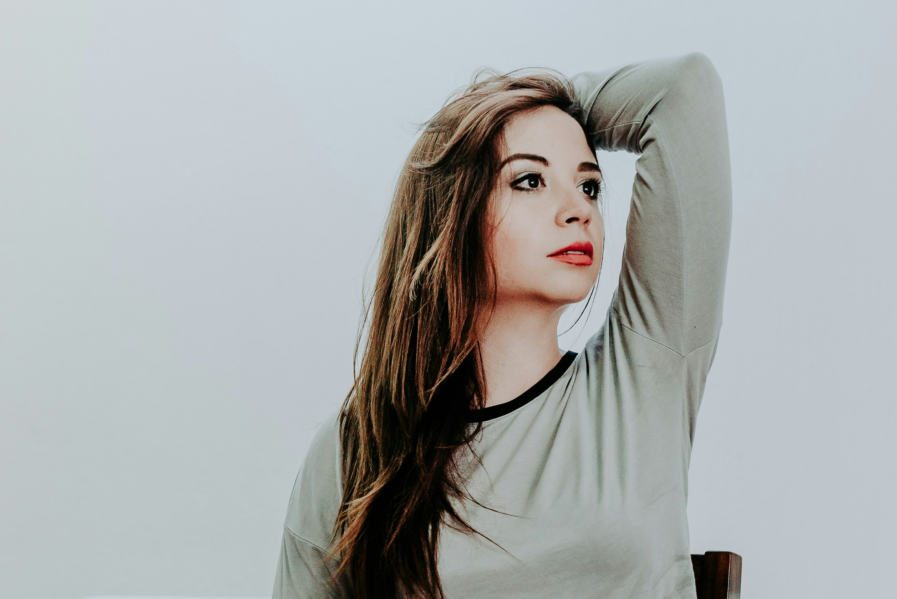 Woman with long hair and casual attire gazes thoughtfully while resting her arm on her head against a minimalist background.