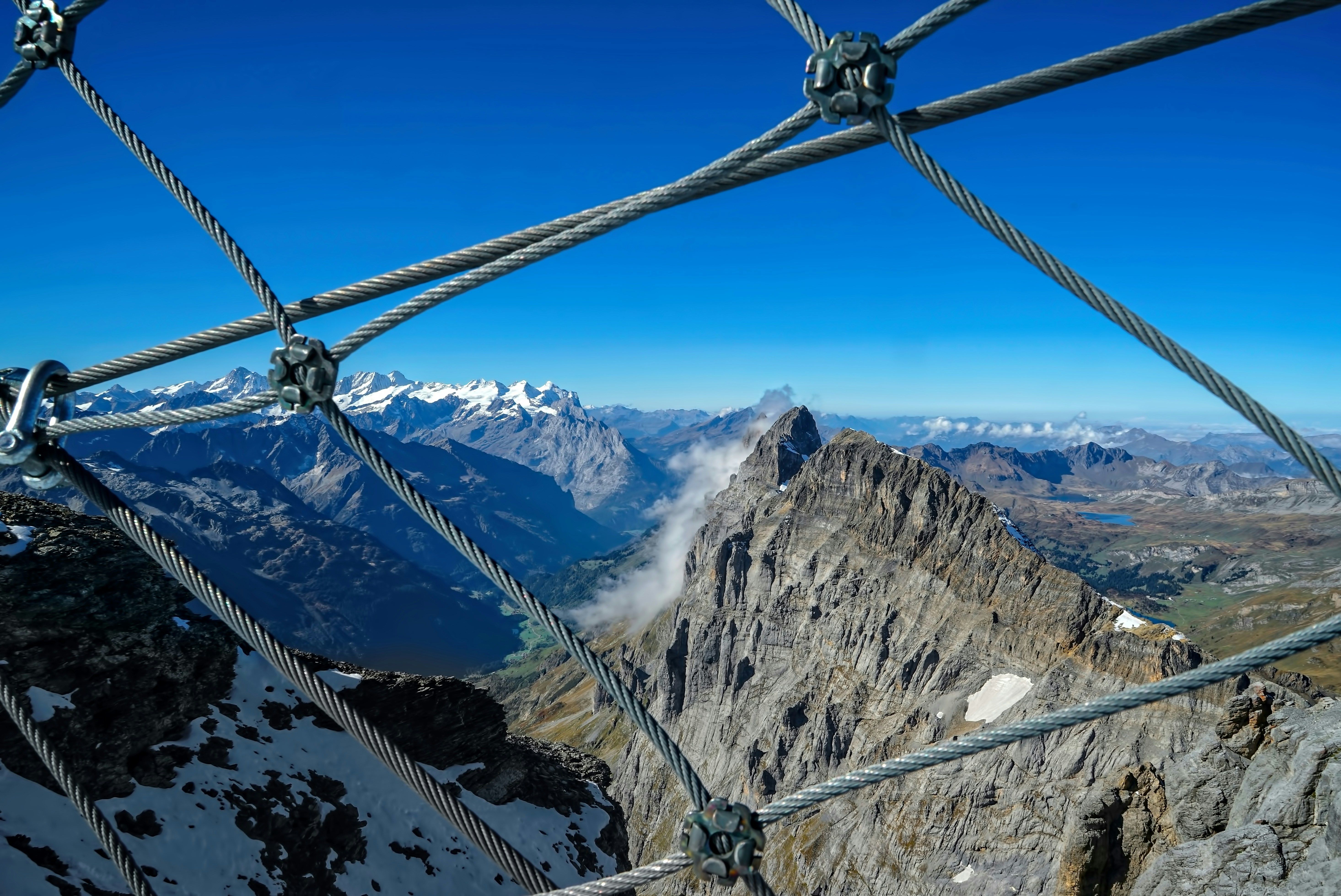 Breathtaking mountain landscape framed by a protective wire mesh, showcasing peaks and valleys under a clear blue sky.
