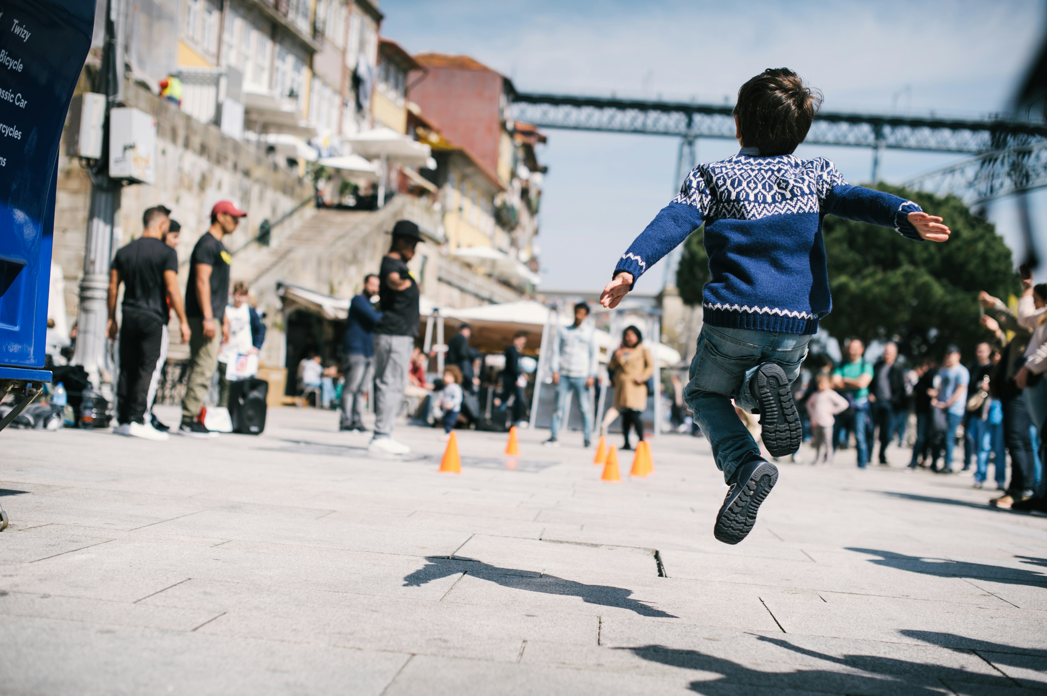 Child in blue outfit mid-jump on a lively city street with people and a bridge in the background.