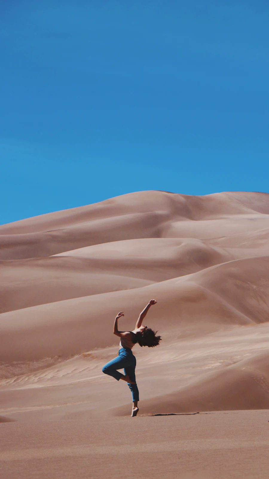 Sand Dancer | woman standing on right foot and raising hands at the bottom of sand dunes during daytime