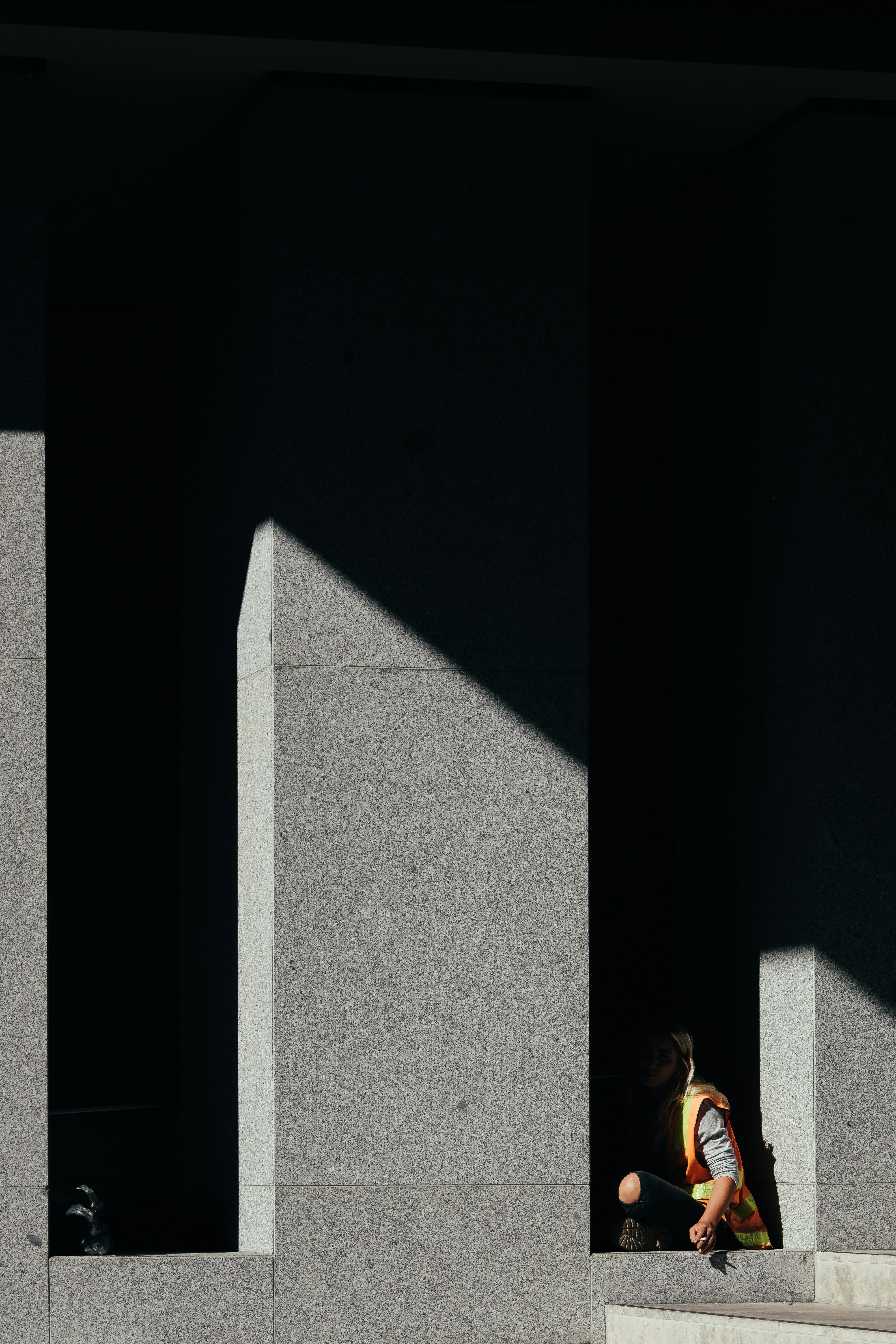 A construction worker in a reflective vest sits amidst towering granite columns, bathed in contrasting shadows. The scene captures the intersection of human presence and architectural grandeur.