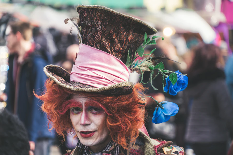 A person dressed as the Mad Hatter wears an oversized patterned top hat with pink ribbon and blue roses. Blurred crowd activity fills the background.