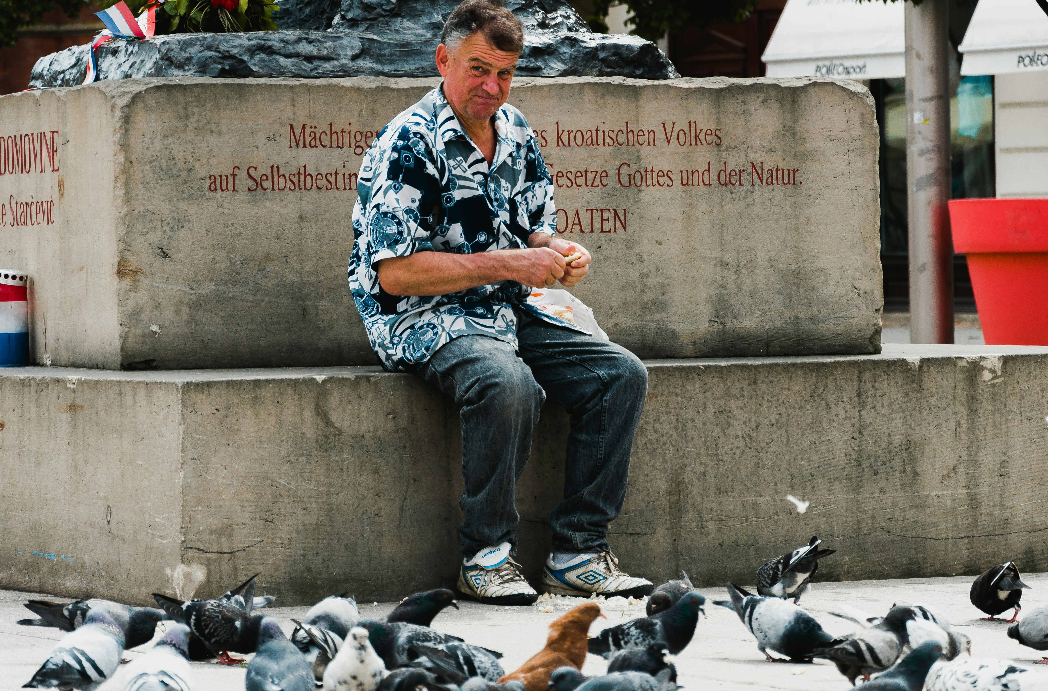 Man seated on a concrete ledge surrounded by pigeons in an urban setting.