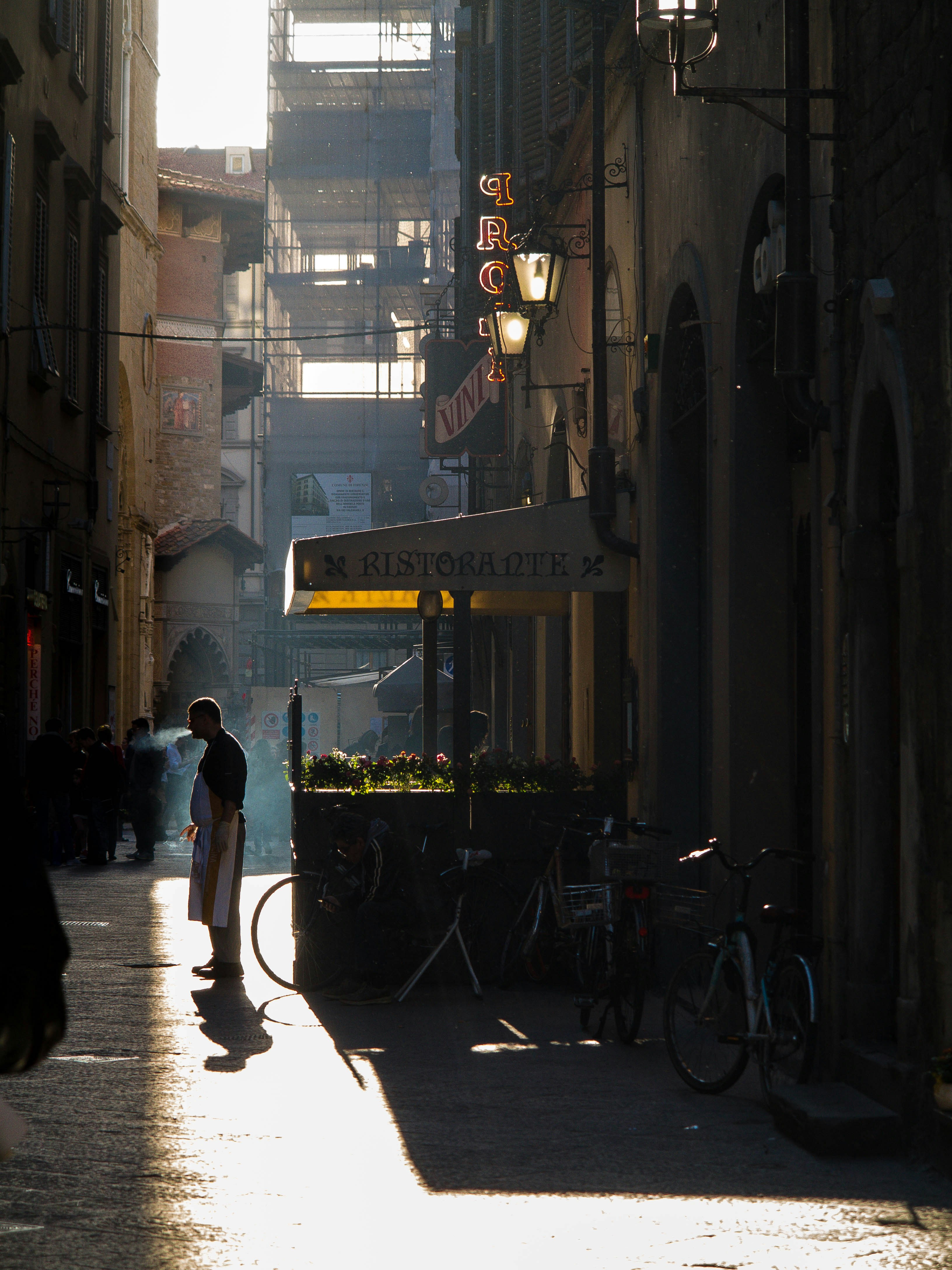 A solitary figure stands outside a quaint restaurant in a narrow alley, bathed in the soft glow of early evening light. The atmosphere is imbued with a sense of anticipation.