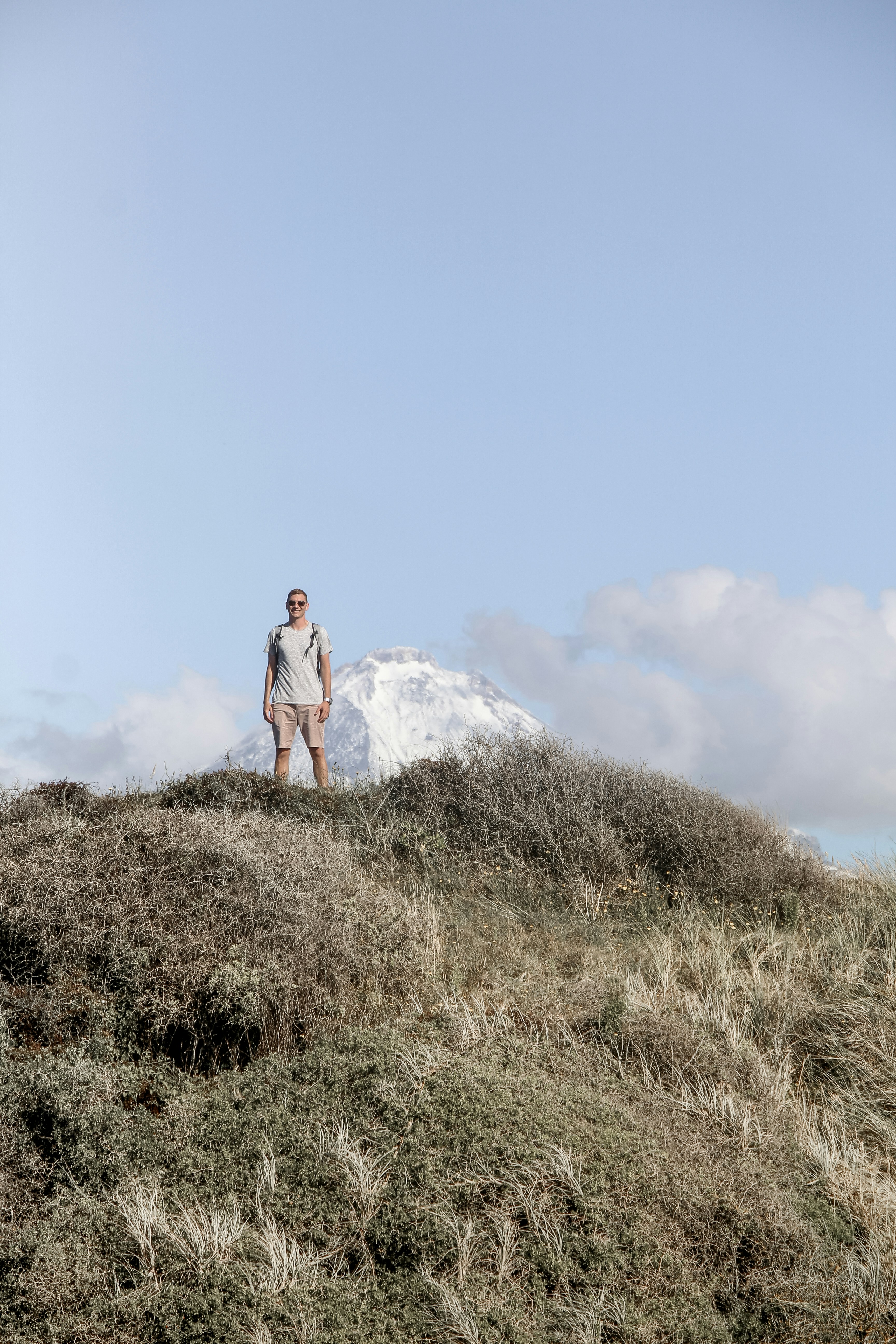 A solitary figure stands atop a grassy hill, with a snow-capped mountain looming in the background under a clear sky.