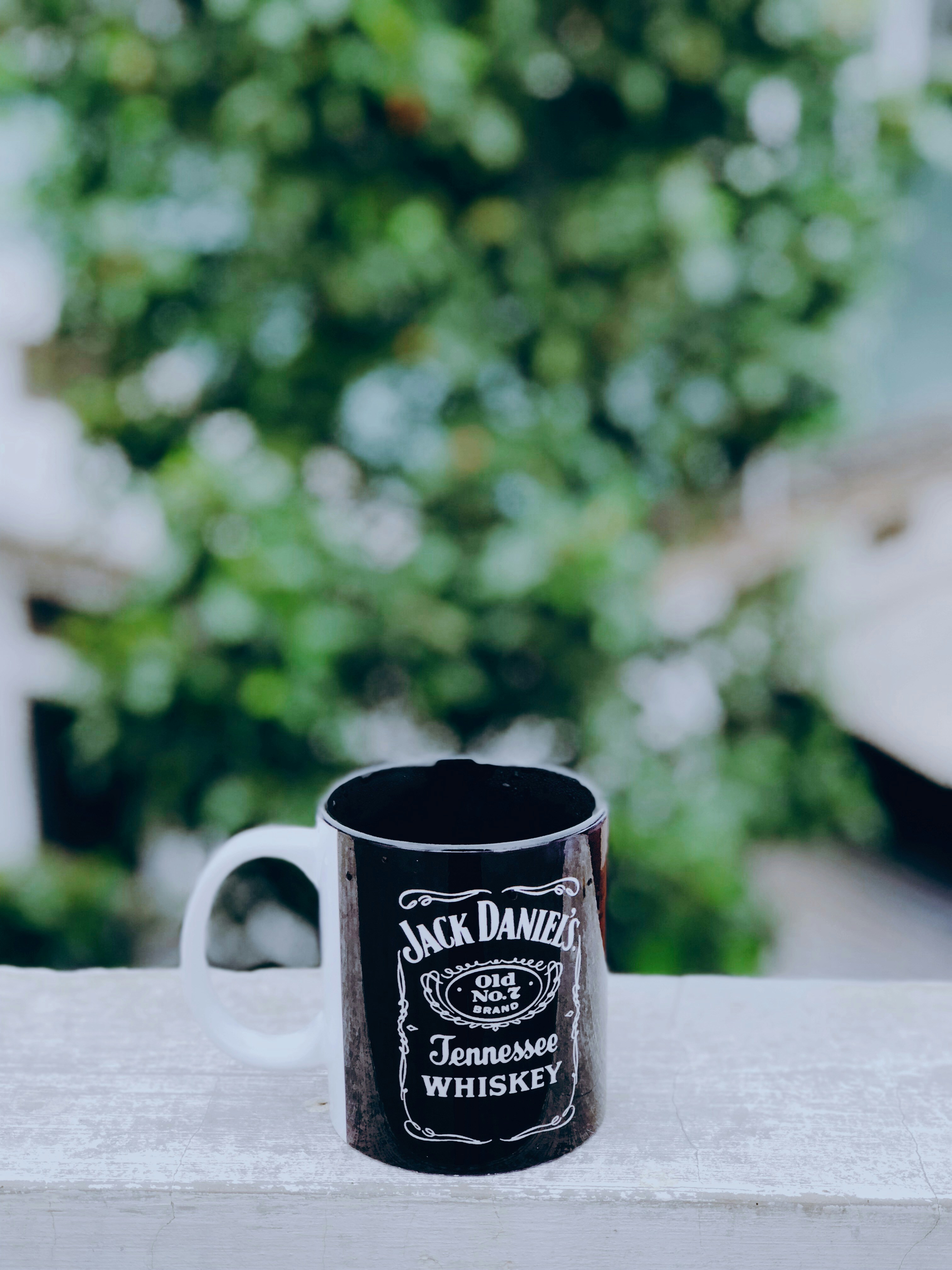Black mug featuring Jack Daniel's logo rests on a railing, with a blurred green backdrop suggesting a tranquil outdoor setting.