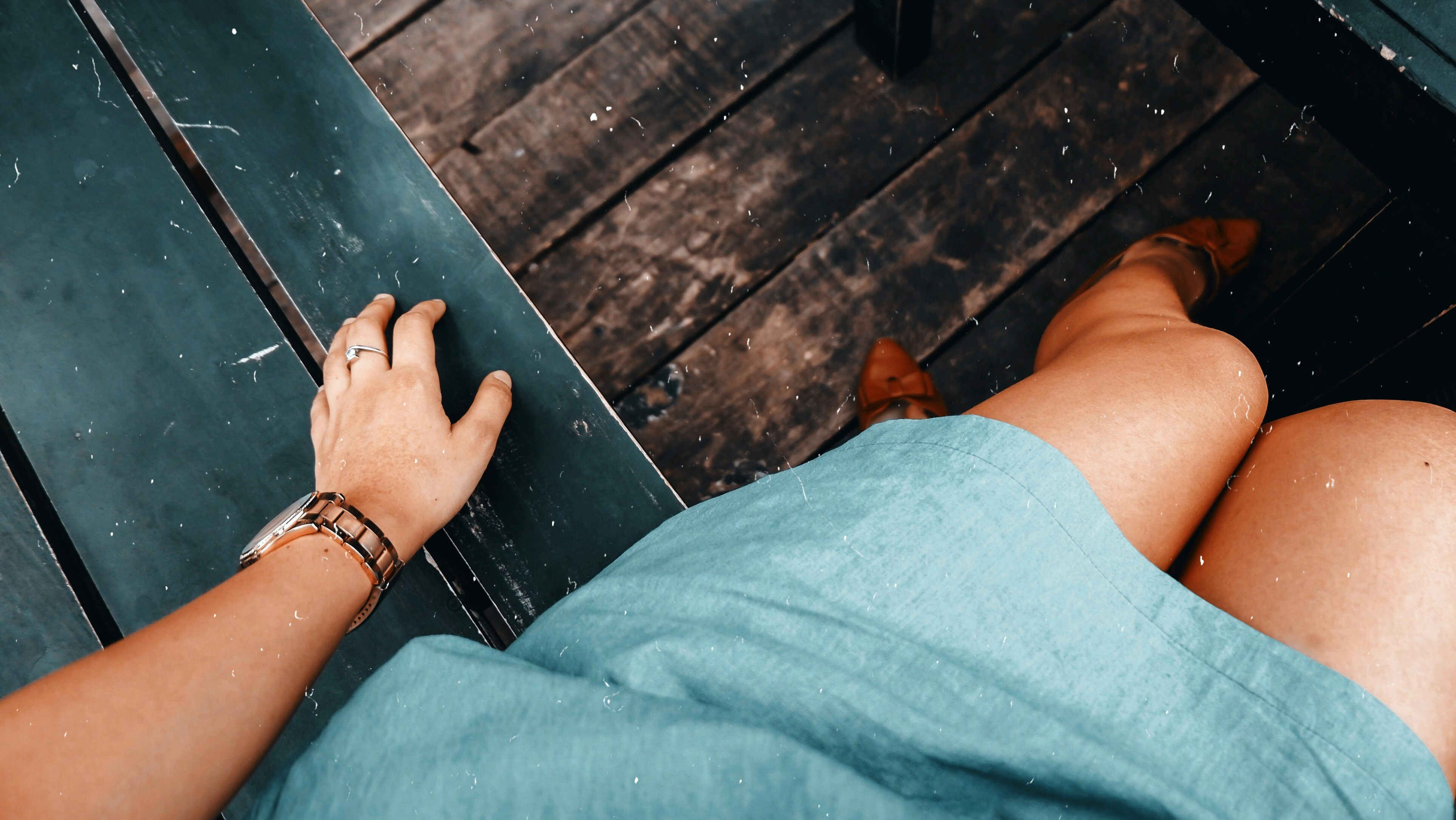 Casual scene featuring a person's hand resting on a wooden bench, with a glimpse of their legs and stylish sandals. The setting evokes a relaxed atmosphere.
