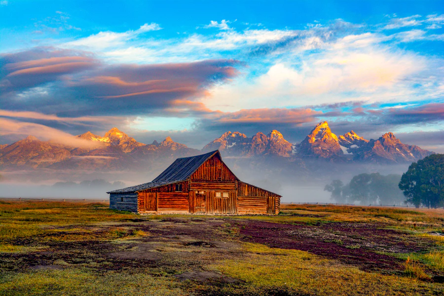 Weathered wooden barn in an open field with misty mountains glowing at sunrise behind it. Dramatic clouds and warm light color the sky above the rugged horizon.