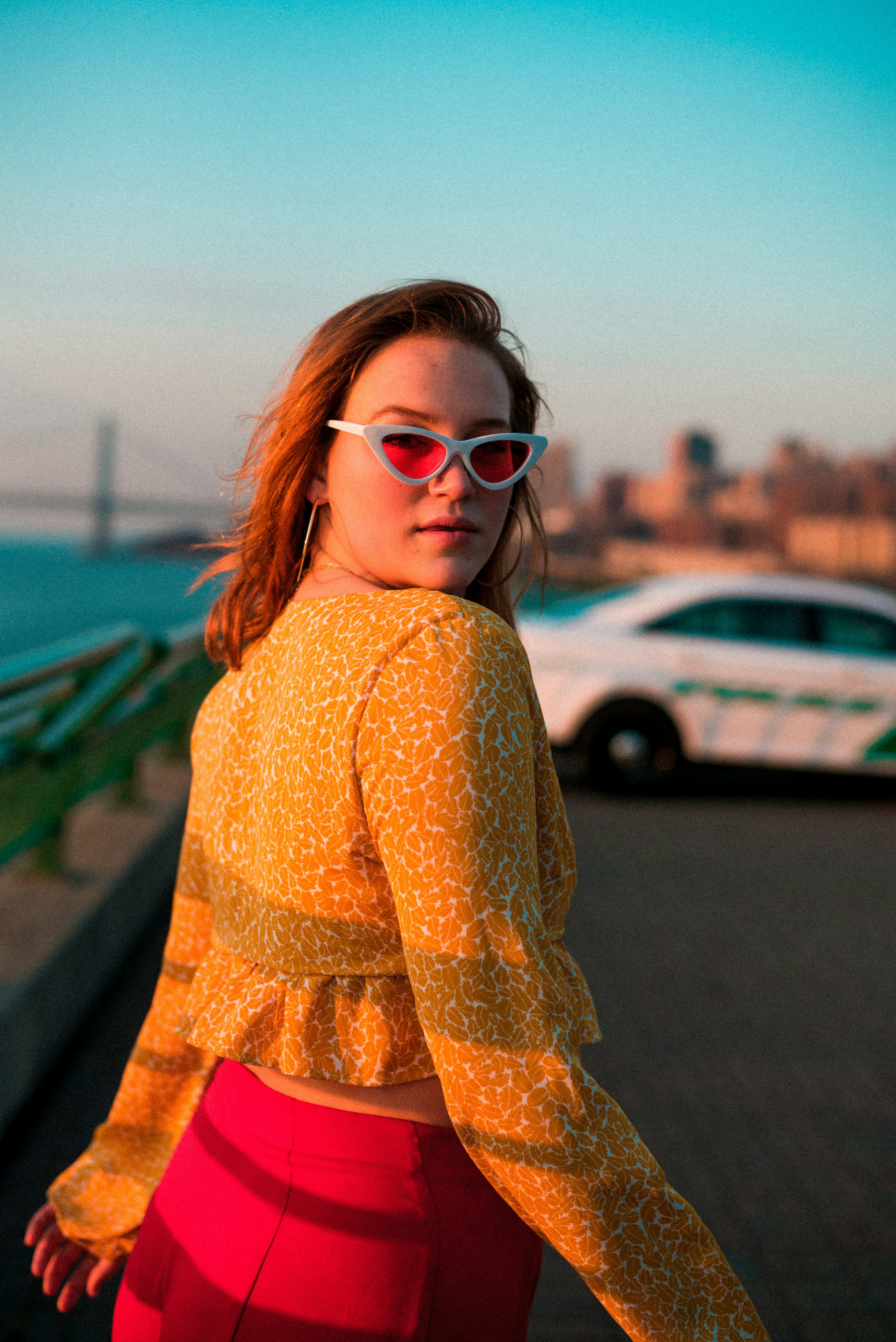 A young woman in a yellow patterned blouse and red pants stands on a waterfront boardwalk at golden hour, wearing white sunglasses. The city skyline and a police car are blurred in the background, giving a warm, cinematic feel. This is a candid lifestyle photograph.