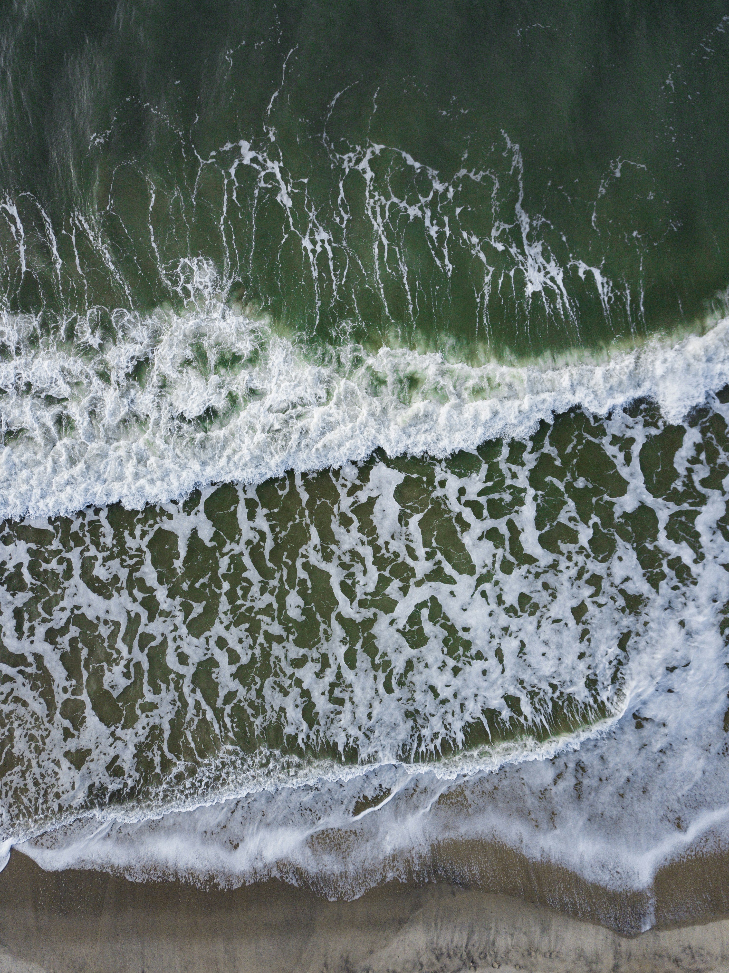 Aerial view of ocean waves crashing onto the sandy beach, showcasing intricate patterns of foam and water. 