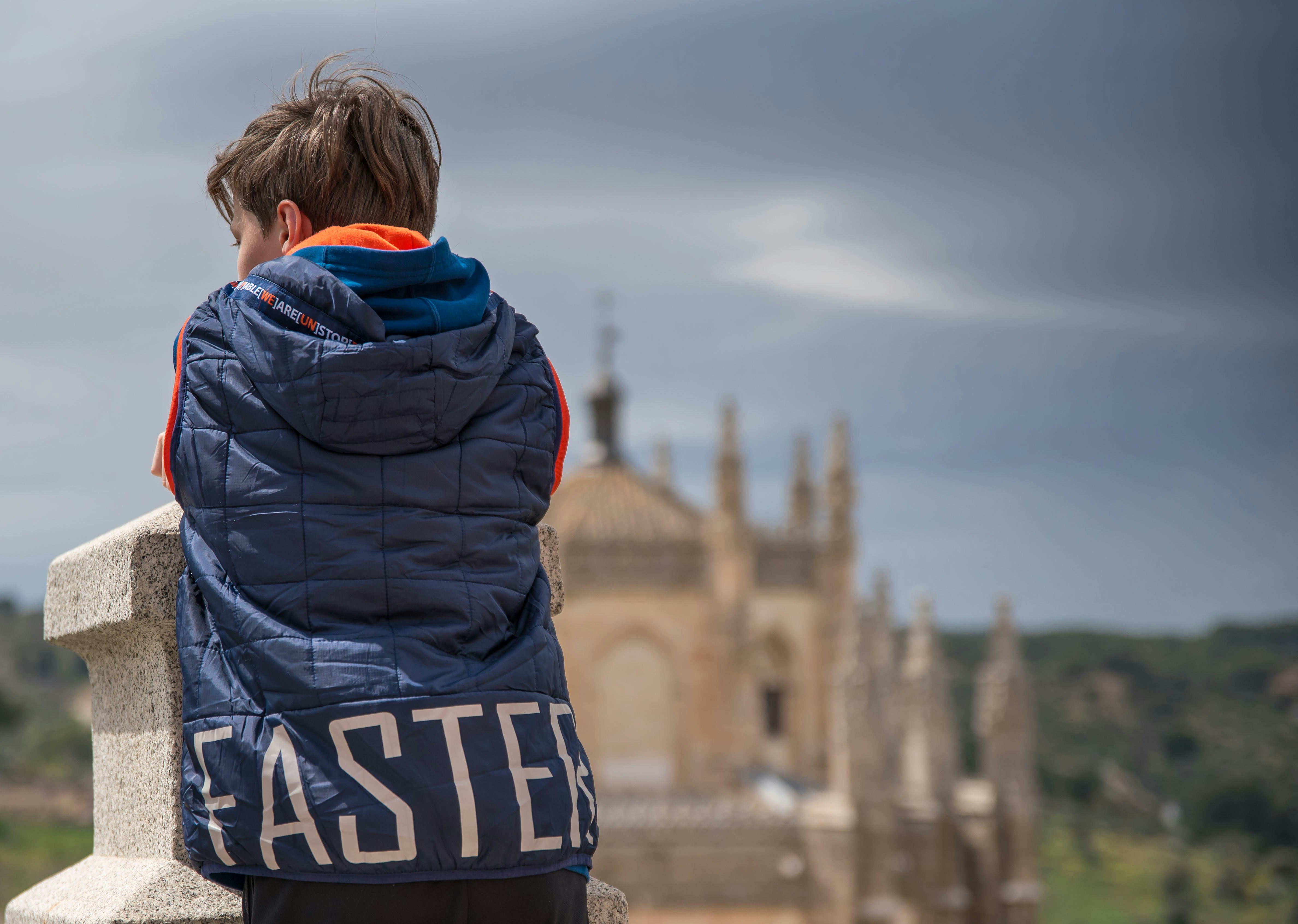 Child in a blue jacket gazing towards an ornate stone building under a cloudy sky.