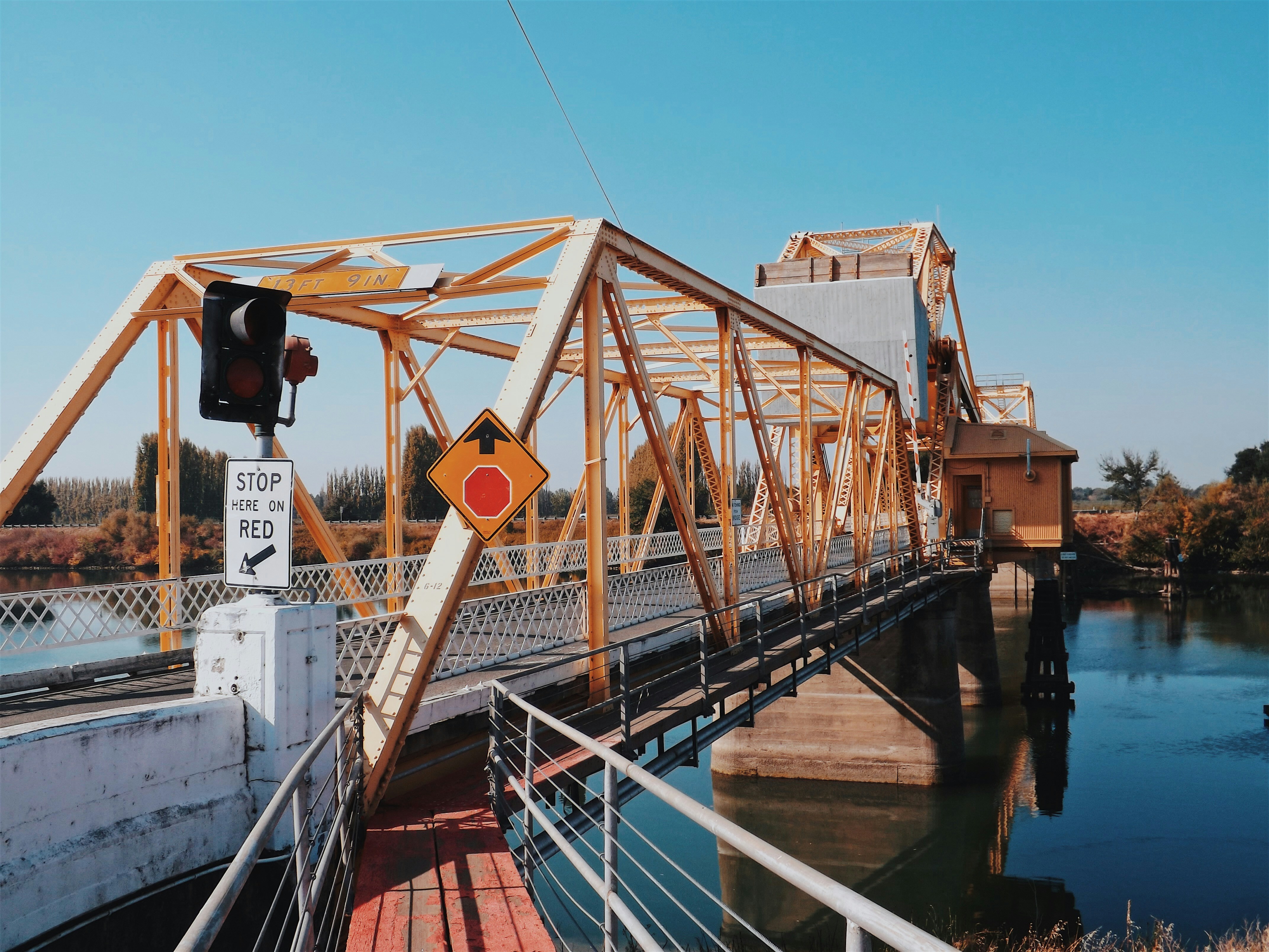 Yellow truss bridge spanning a calm river under a clear blue sky.