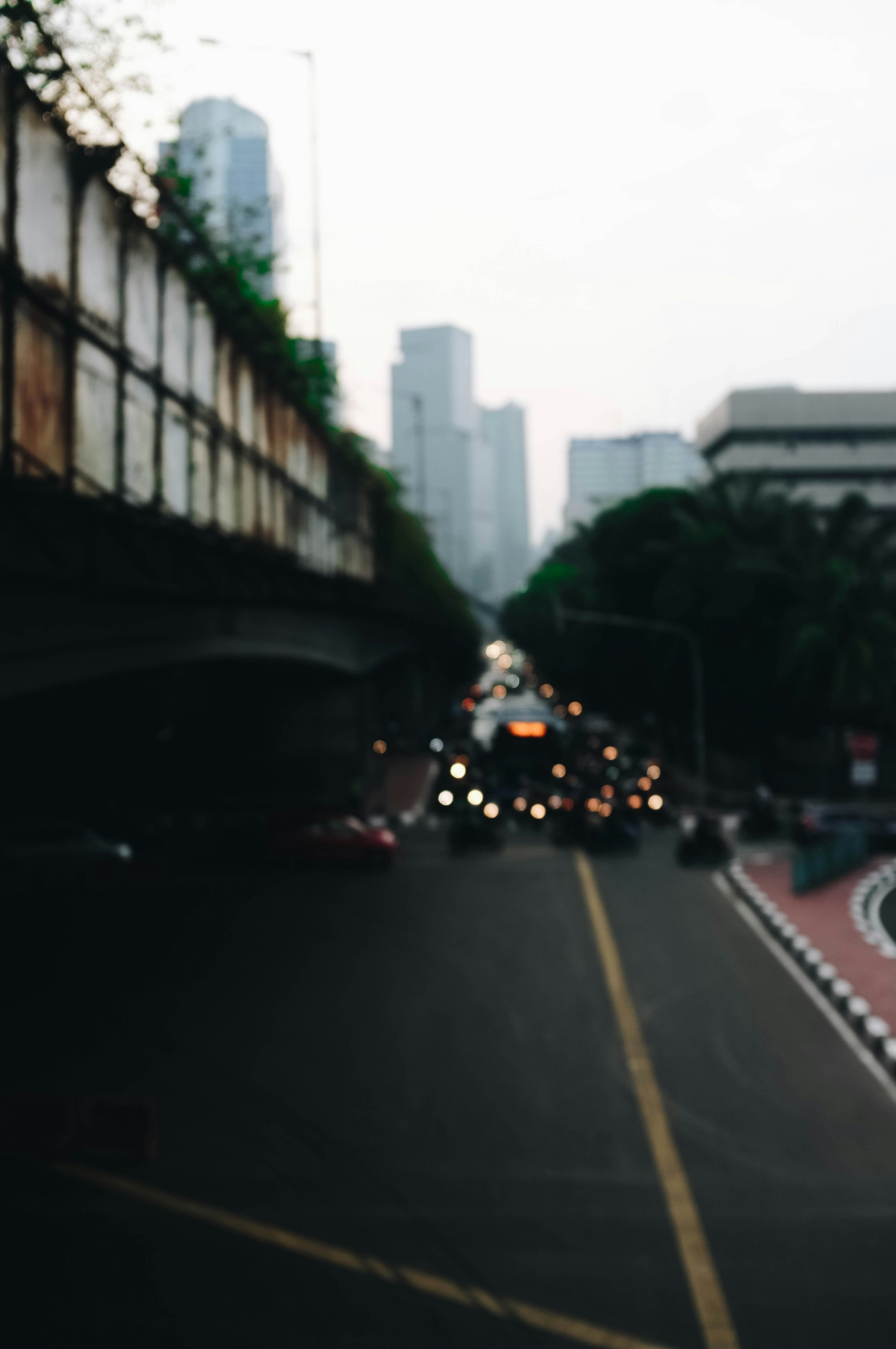 Blurry view of a bustling city street filled with traffic, framed by towering buildings and lush greenery. The scene captures the dynamic rhythm of urban life.