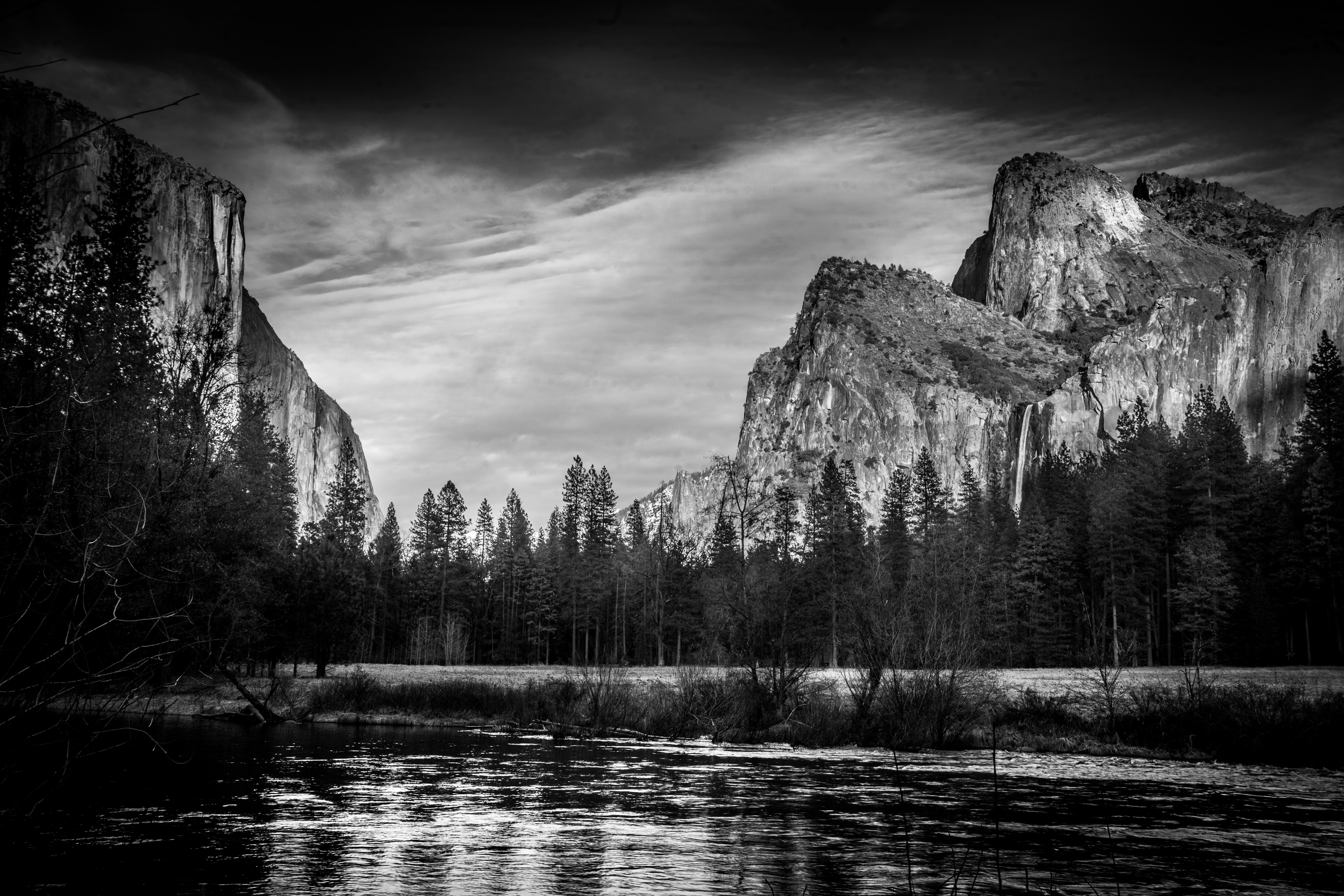 Black and white view of Yosemite Valley with towering granite cliffs and a flowing river under dramatic clouds.