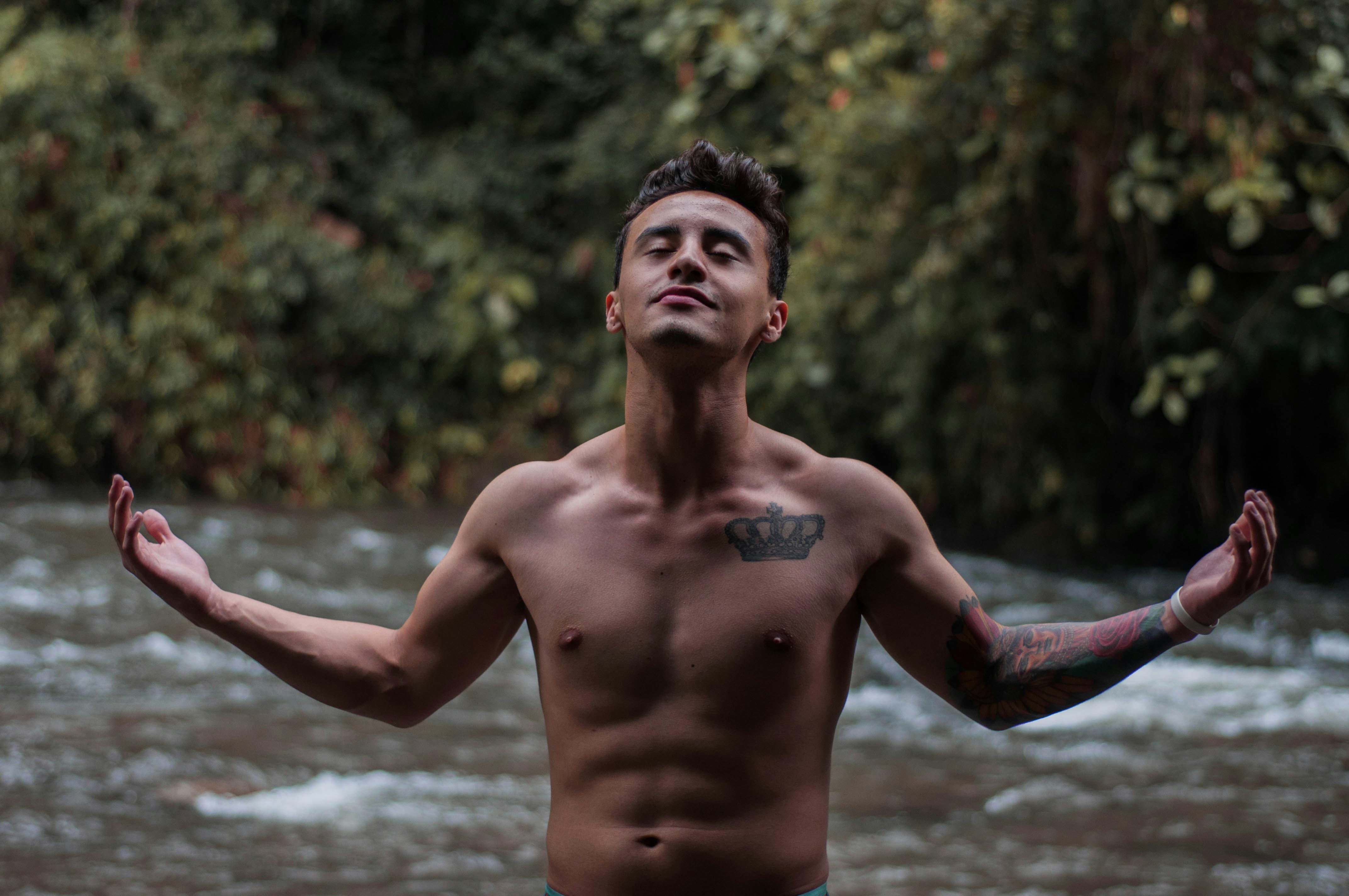 Shirtless man with closed eyes stands with arms outstretched by a flowing river, surrounded by lush greenery.