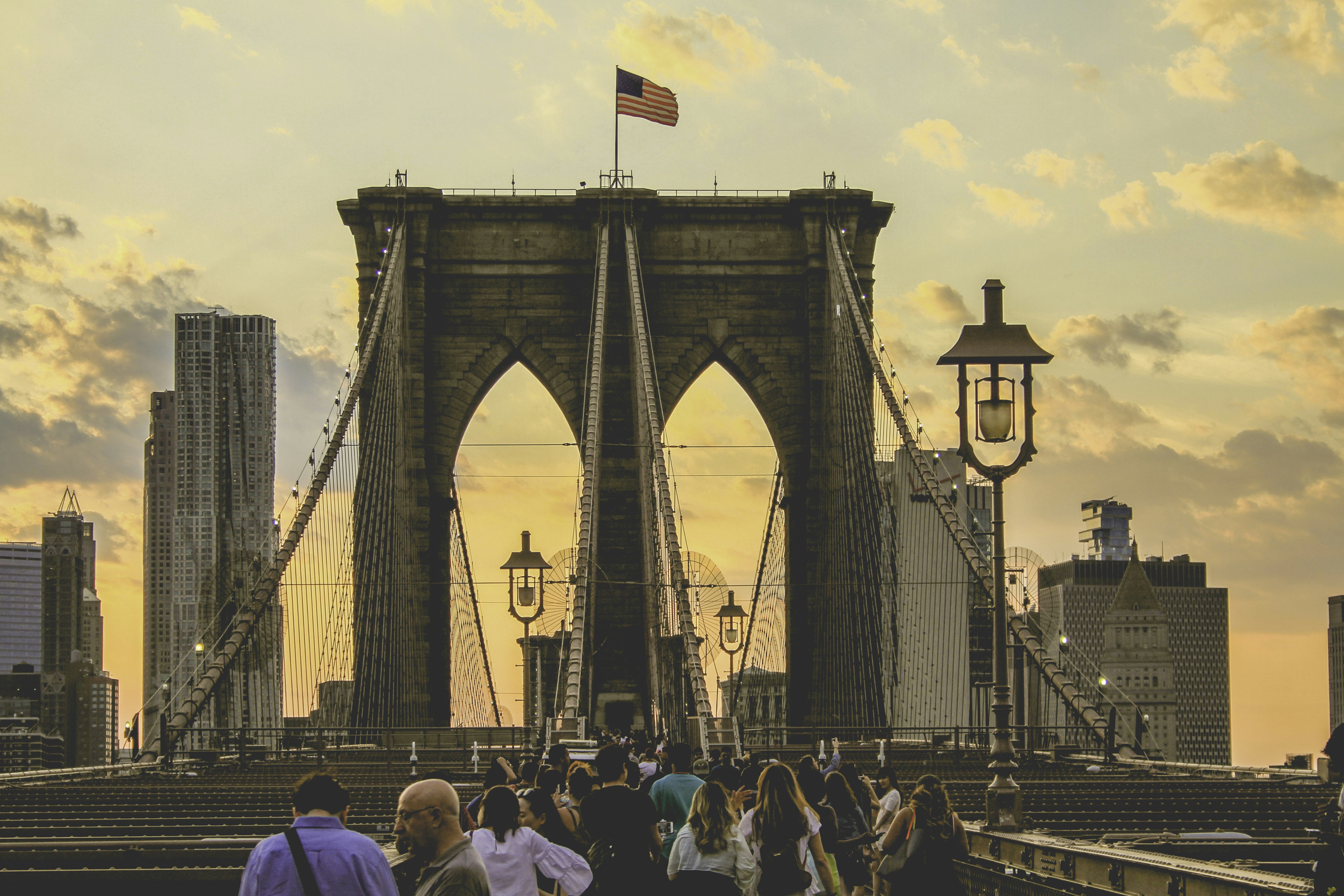 Pedestrians walking on the Brooklyn Bridge at sunset with the city skyline in the background.