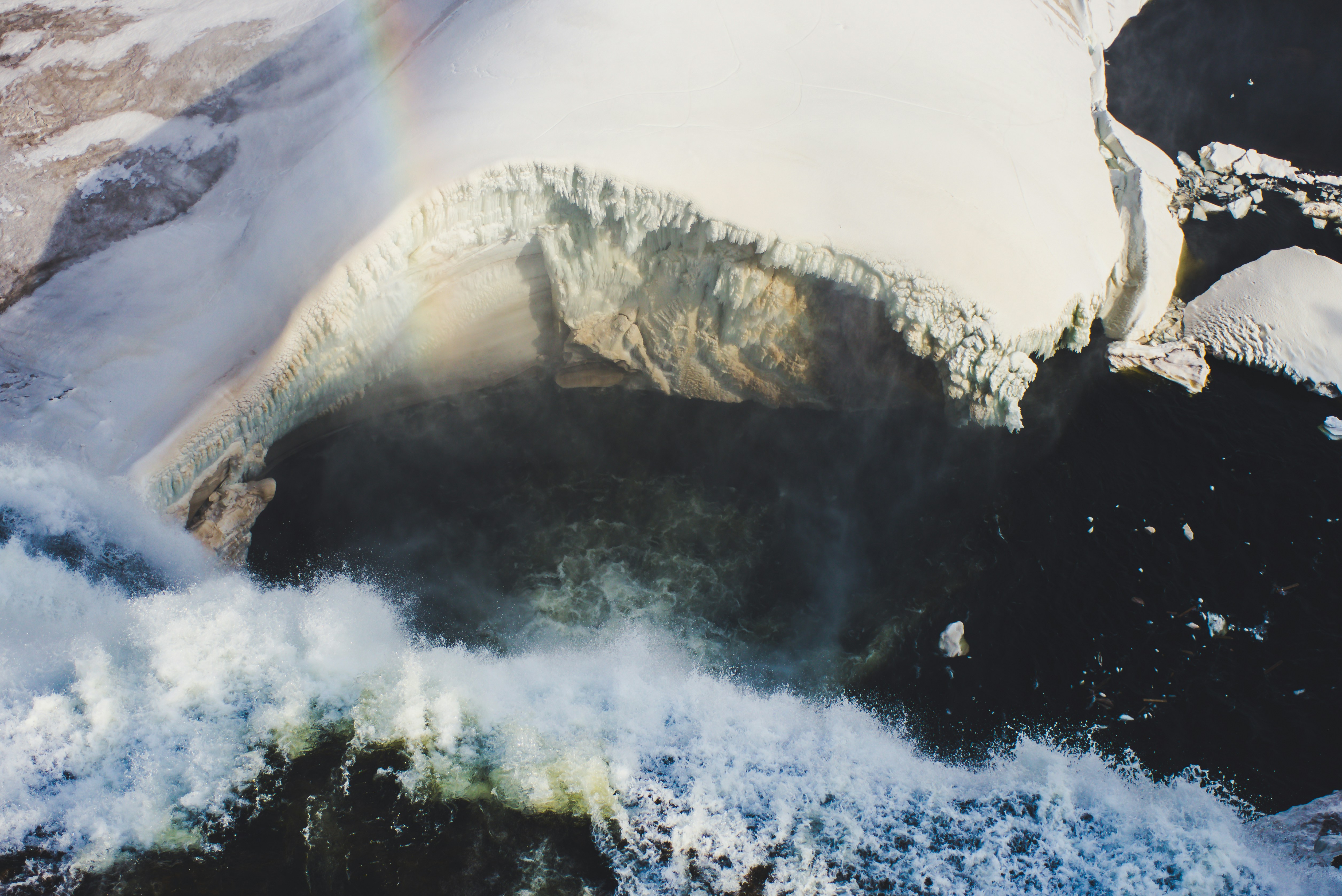 A waterfall plunges into a dark pool surrounded by snow and ice, with a faint rainbow arcing over the scene.