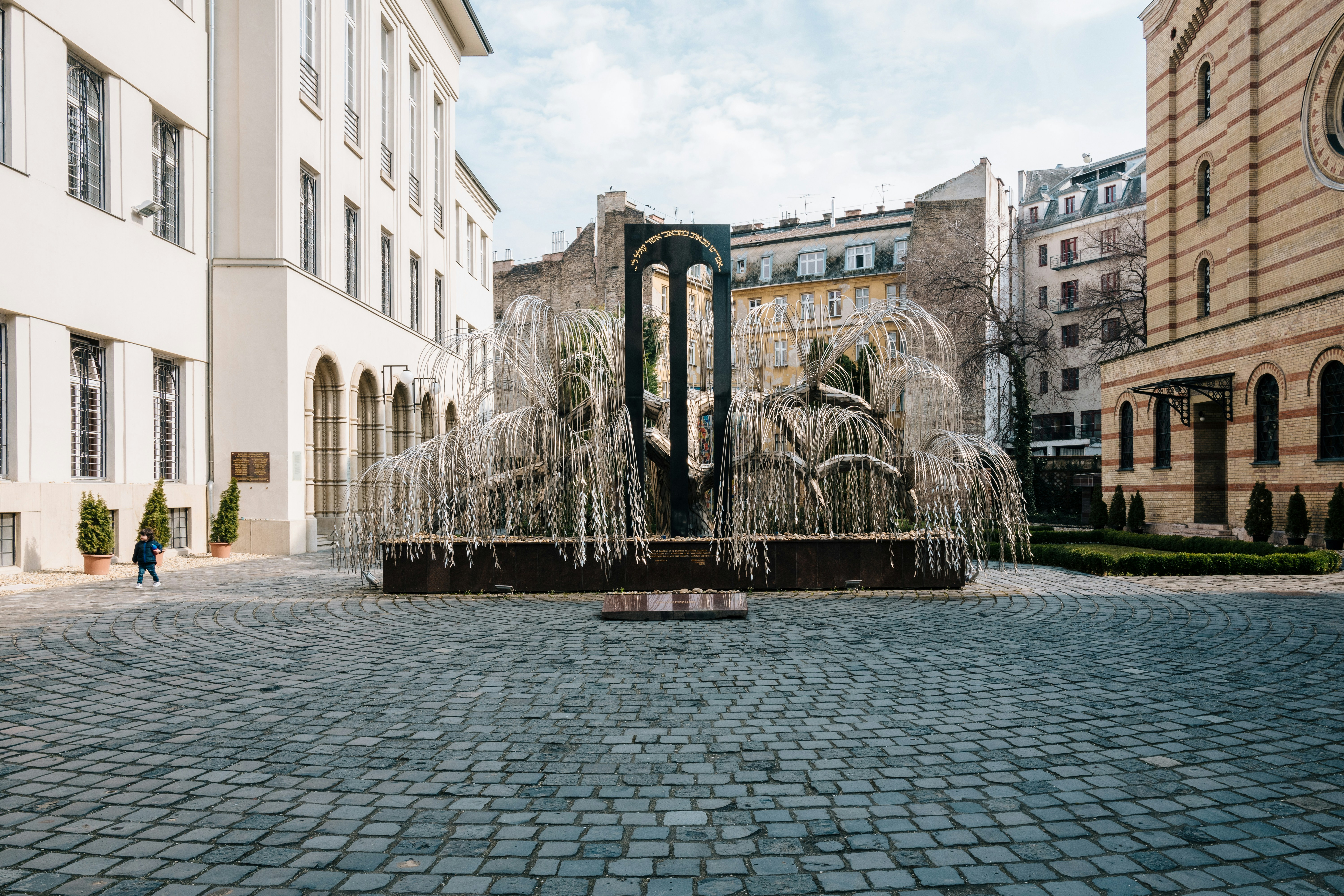 This image captures a tranquil courtyard featuring a striking metal sculpture resembling a weeping willow. The composition is centered, with cobblestone paving leading the eye towards the artwork, framed by the surrounding architecture. The scene is bathed in soft natural light, highlighting the contrast between the sculpture's silvery tones and the warm hues of the buildings, creating a calm and contemplative atmosphere.