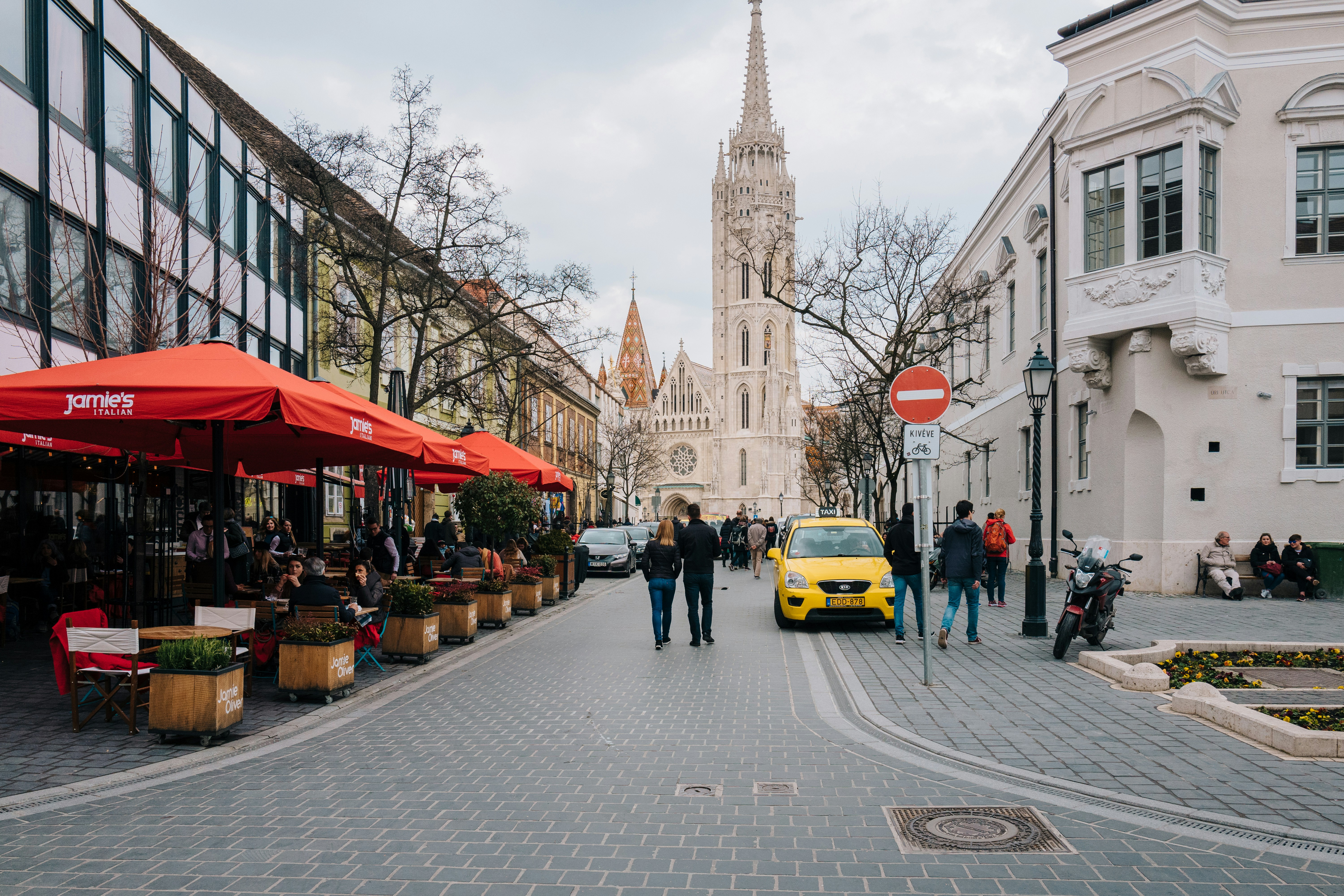 Bustling street scene with pedestrians and a yellow car approaching a towering cathedral under a cloudy sky.