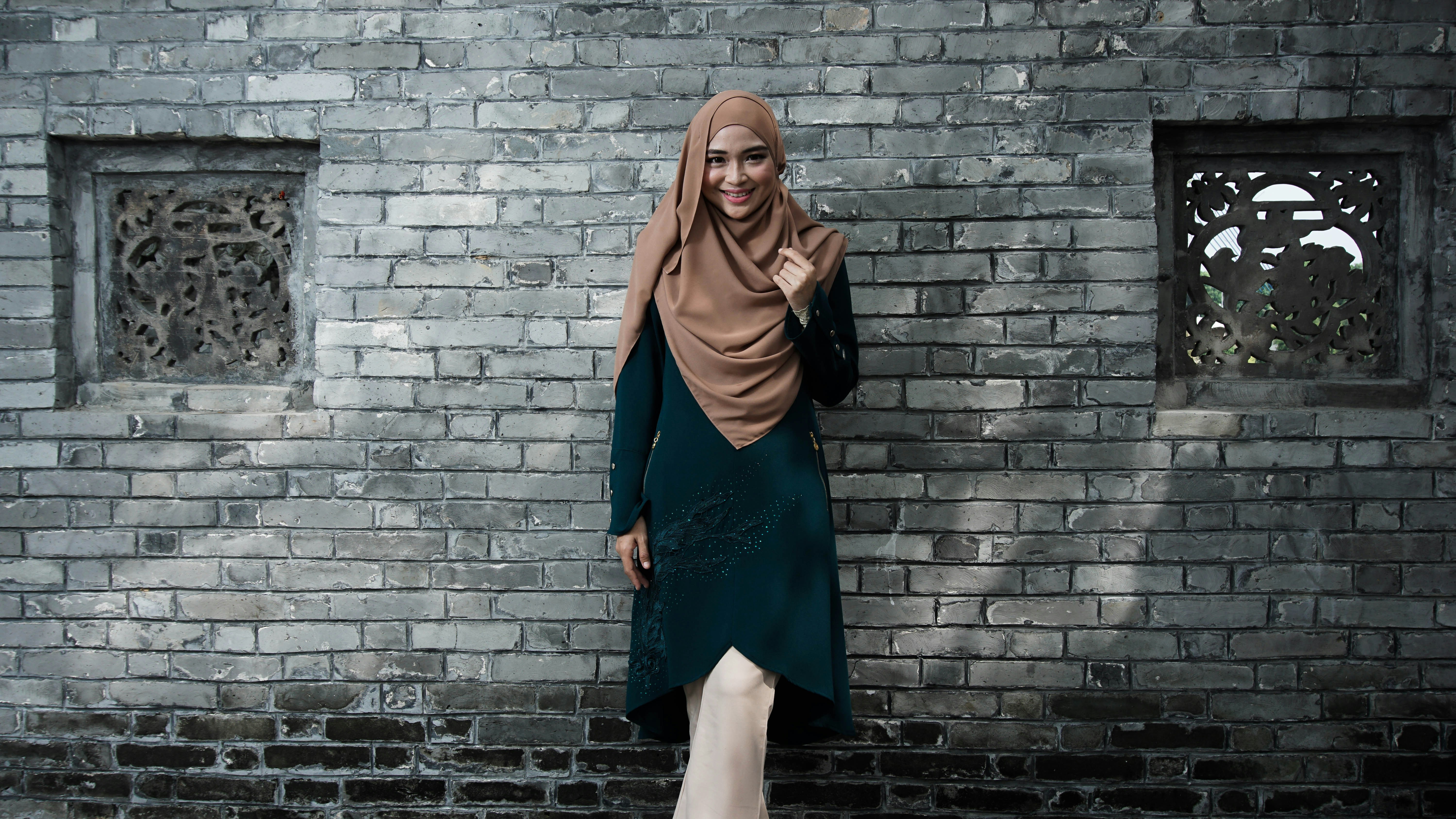 Woman in a hijab standing against a textured brick wall with decorative insets.