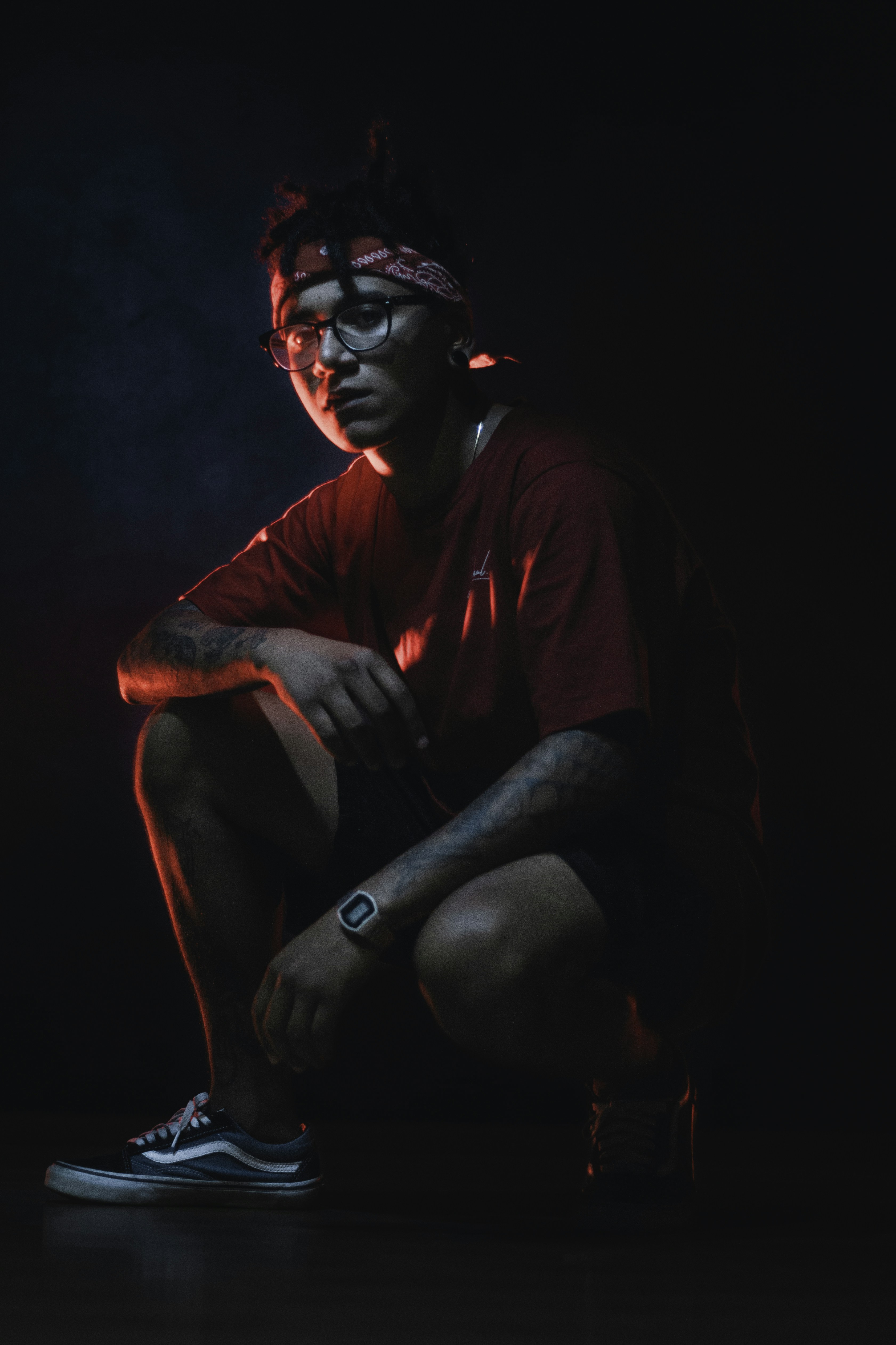 Person in a red t-shirt and black shorts kneeling in dramatic low lighting with a bandana and glasses.