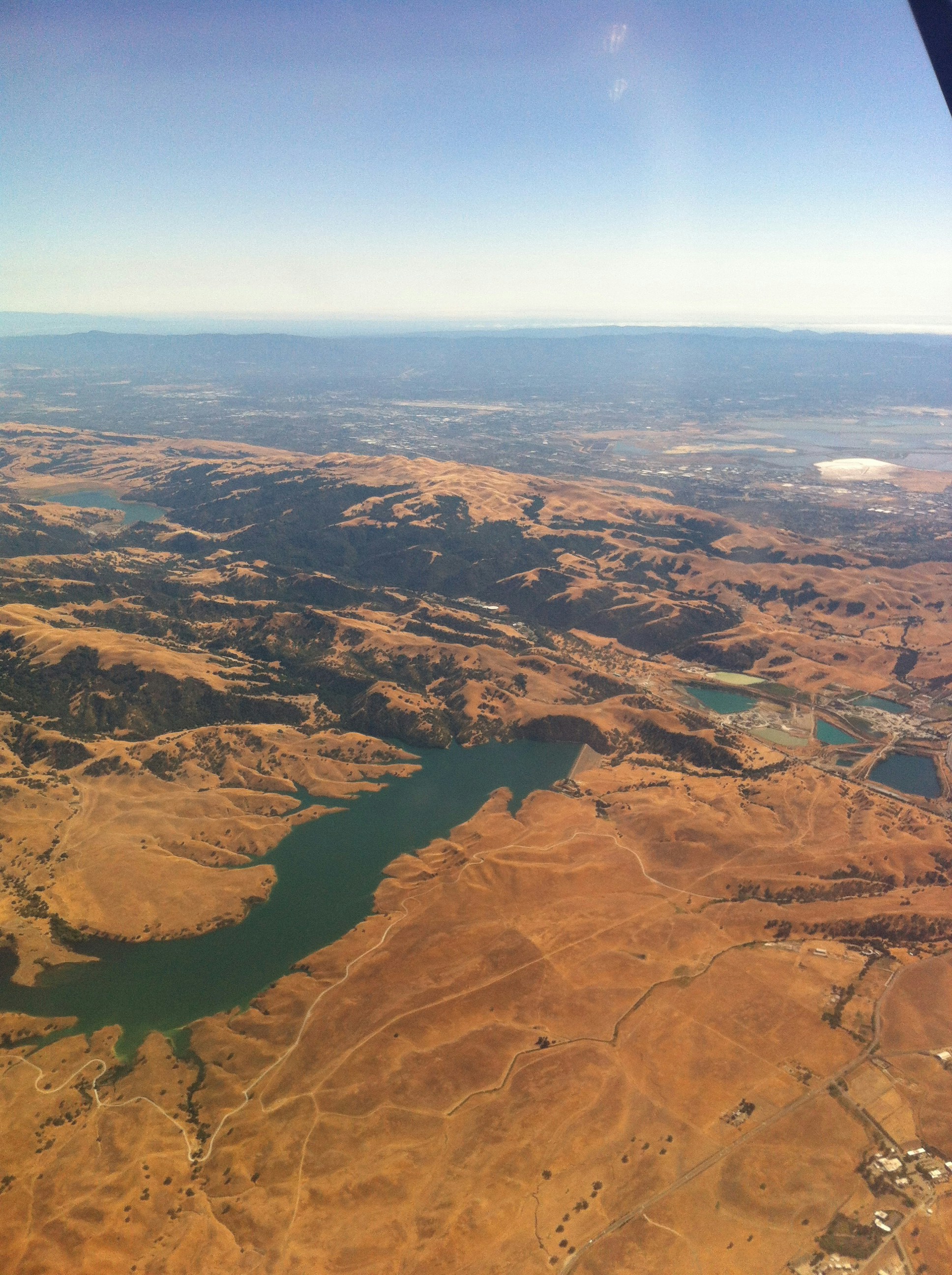 Aerial view of arid canyons and winding water bodies under a clear blue sky. The scene emphasizes the earth-toned terrain and distant horizon.
