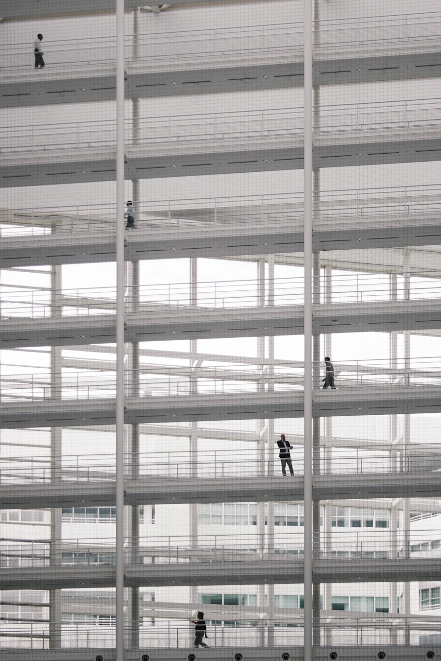 Several small figures walk across stacked white pedestrian bridges inside a bright modern atrium, seen through a fine mesh or grid. The repeating lines and vertical supports create a layered architectural pattern.