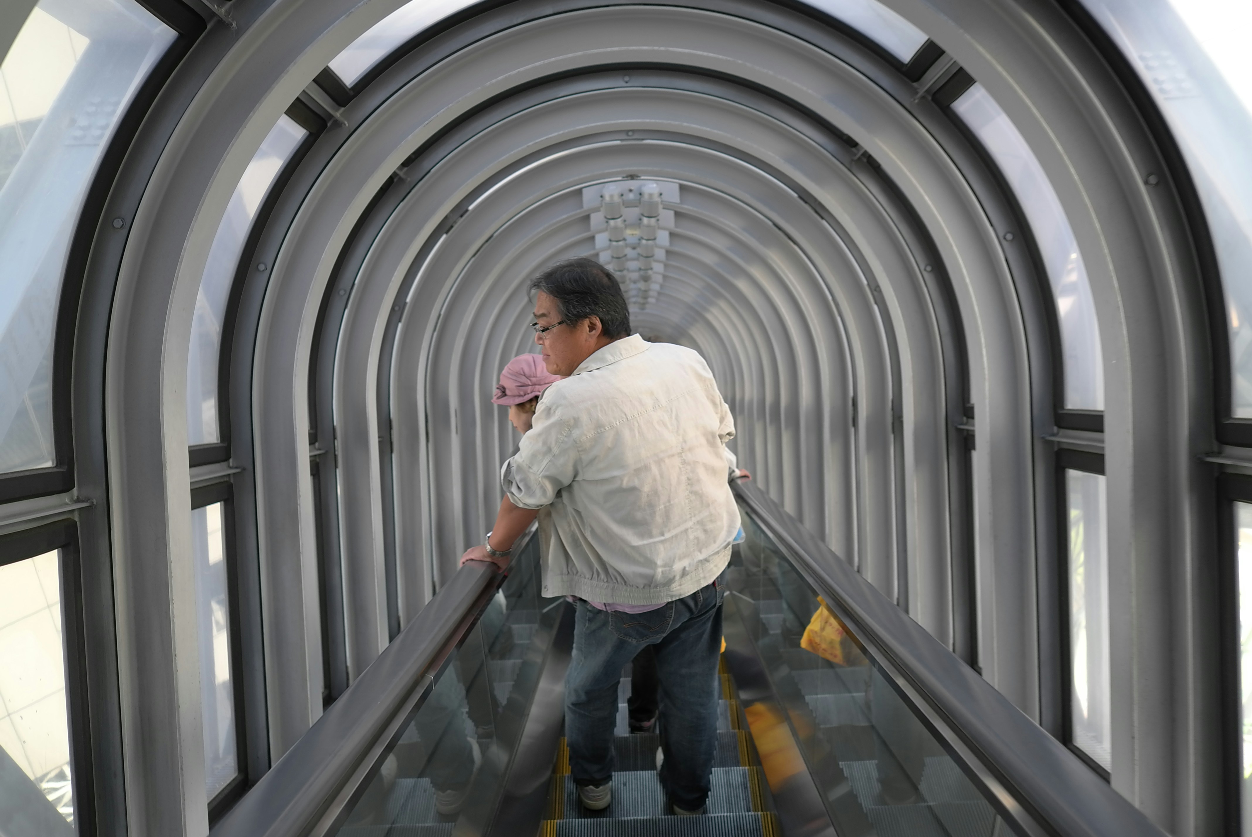 A man and child navigate an enclosed glass tunnel on an escalator. The curved metal arches frame a bright corridor ahead.