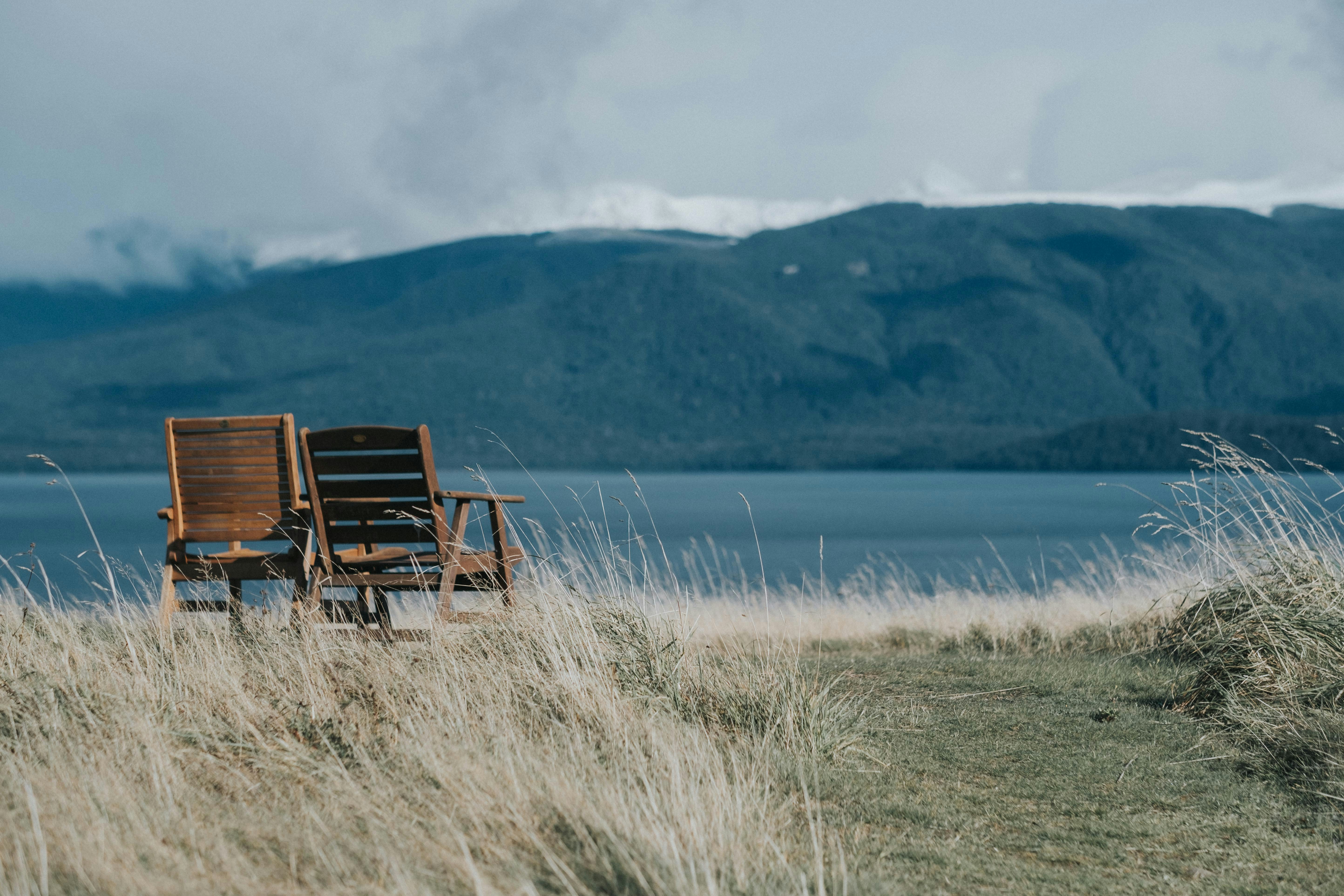 Two wooden chairs facing a tranquil lake with distant mountains under a cloudy sky.