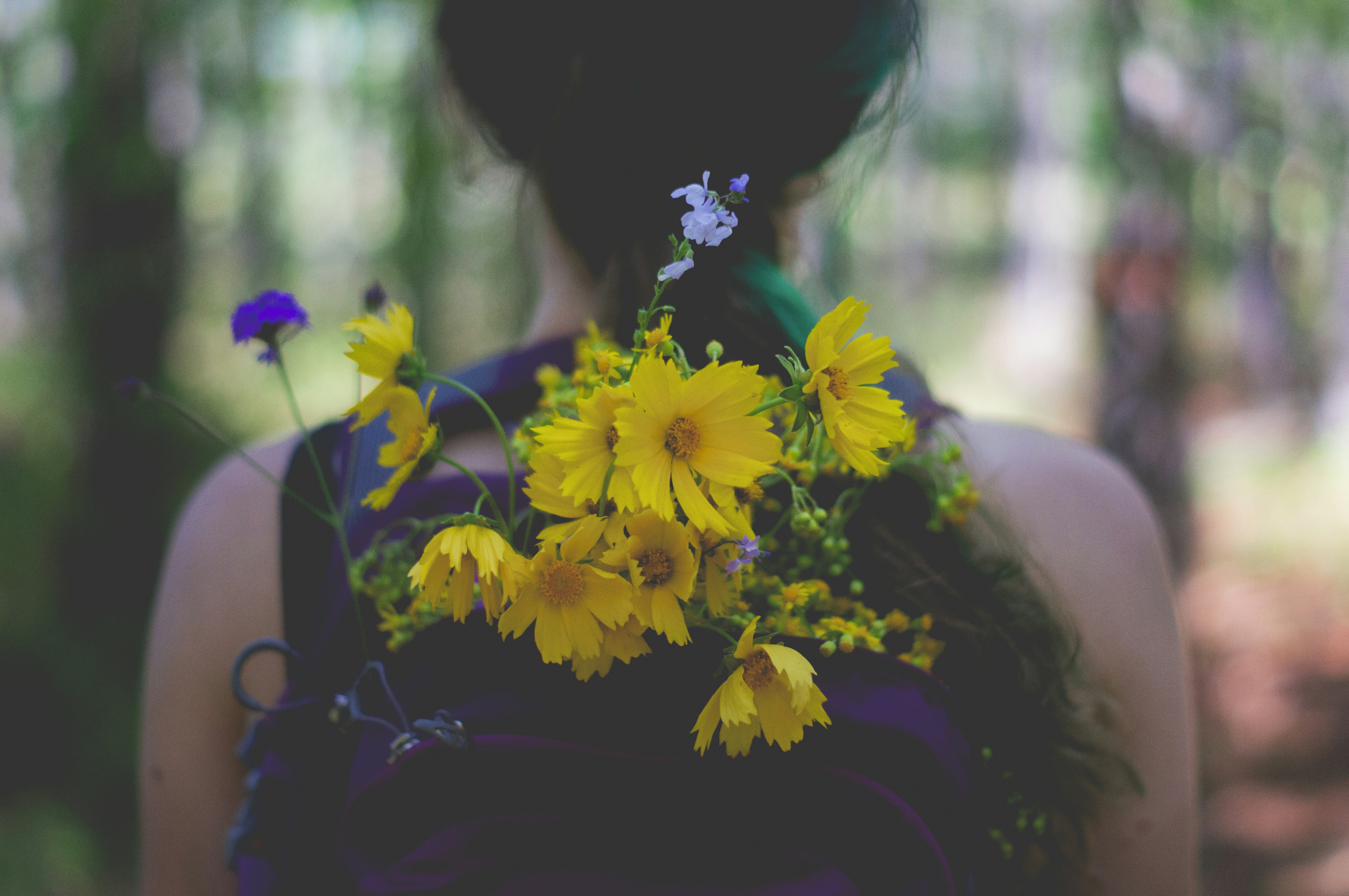 A person with a backpack adorned with vibrant yellow flowers walks through a lush forest, blending nature with personal style.