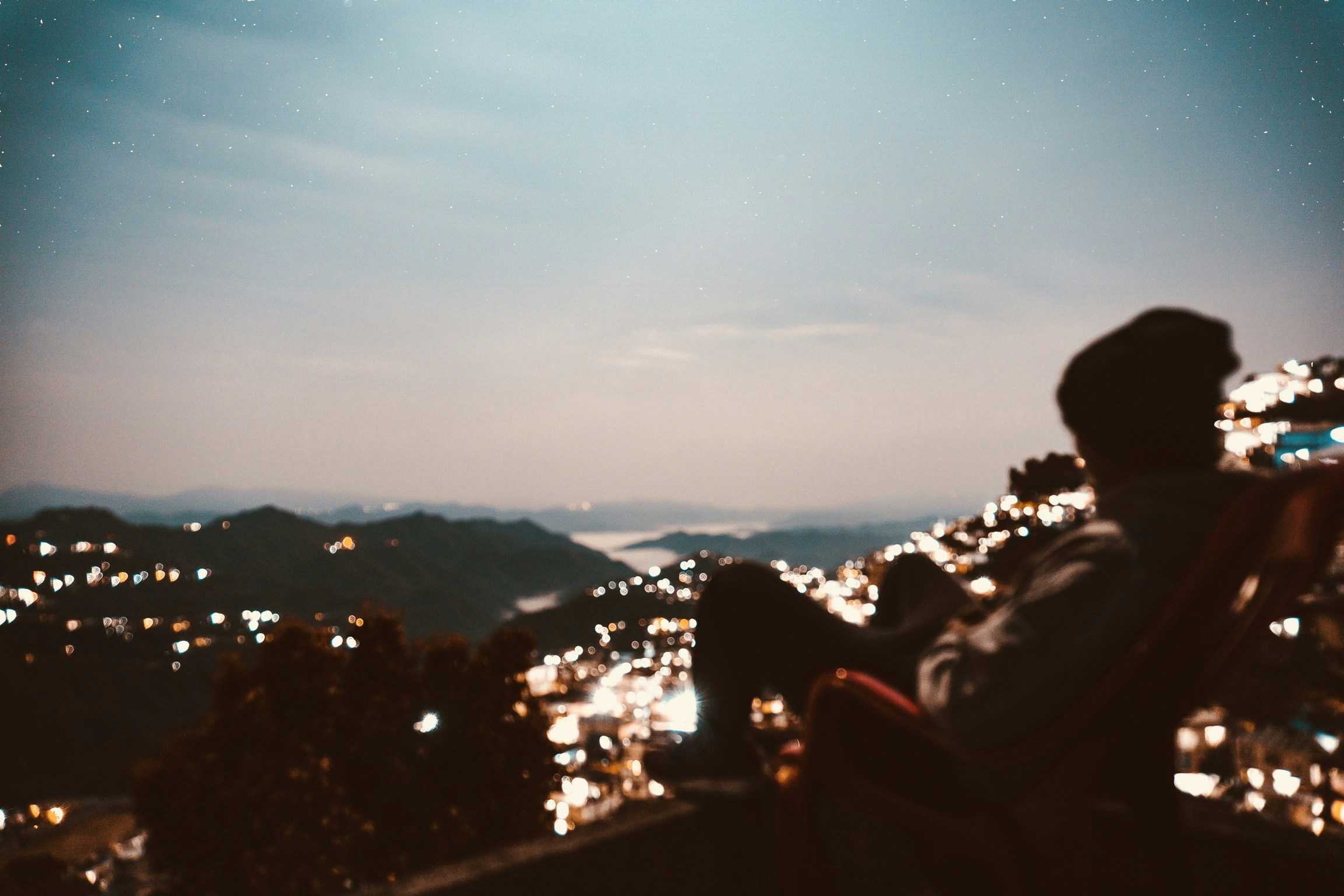 Silhouette of a person sitting on a ledge, overlooking a cityscape illuminated by scattered lights under a twilight sky.