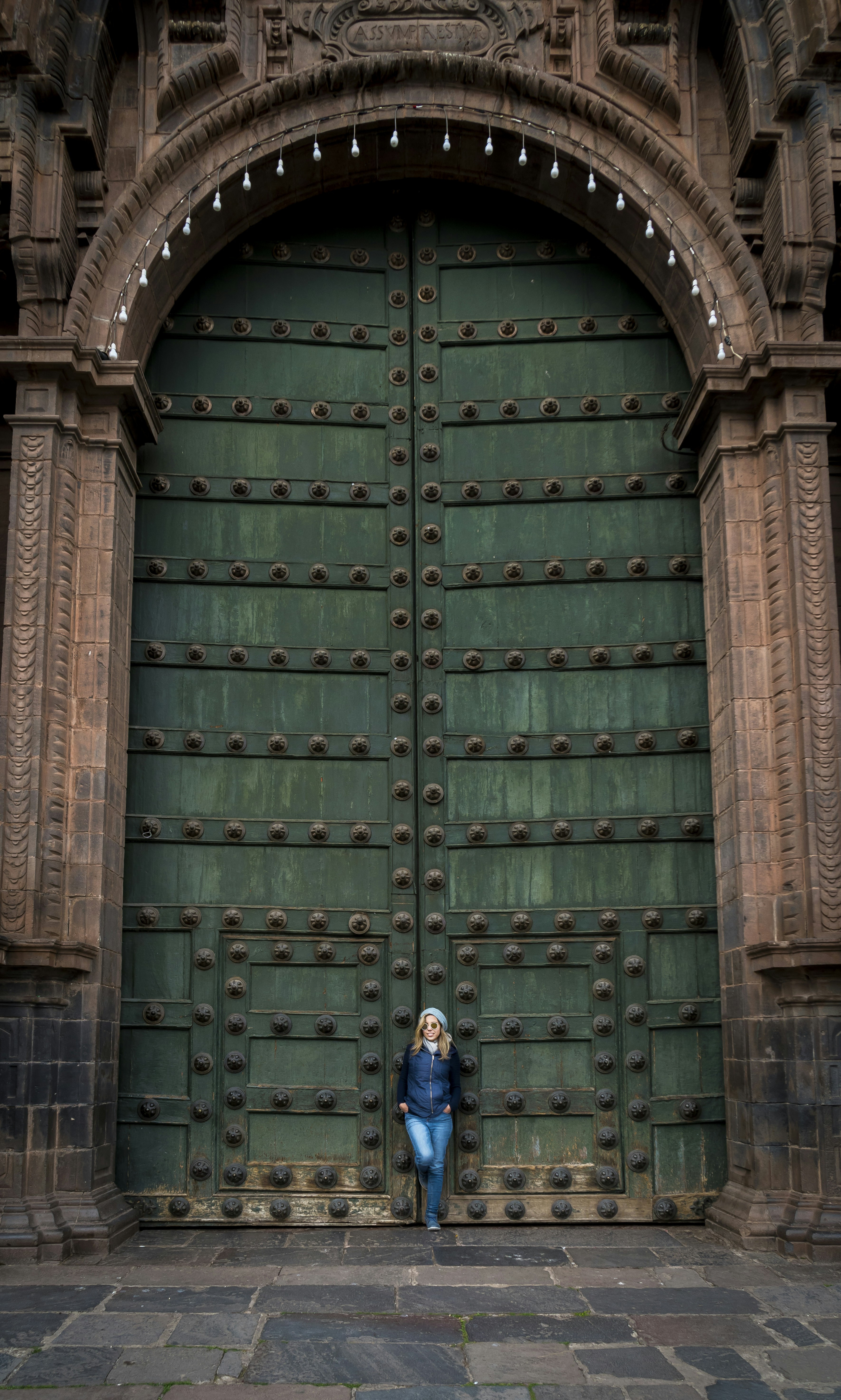 A woman stands beside an enormous, intricately designed green door adorned with metal studs, set against a textured stone wall.