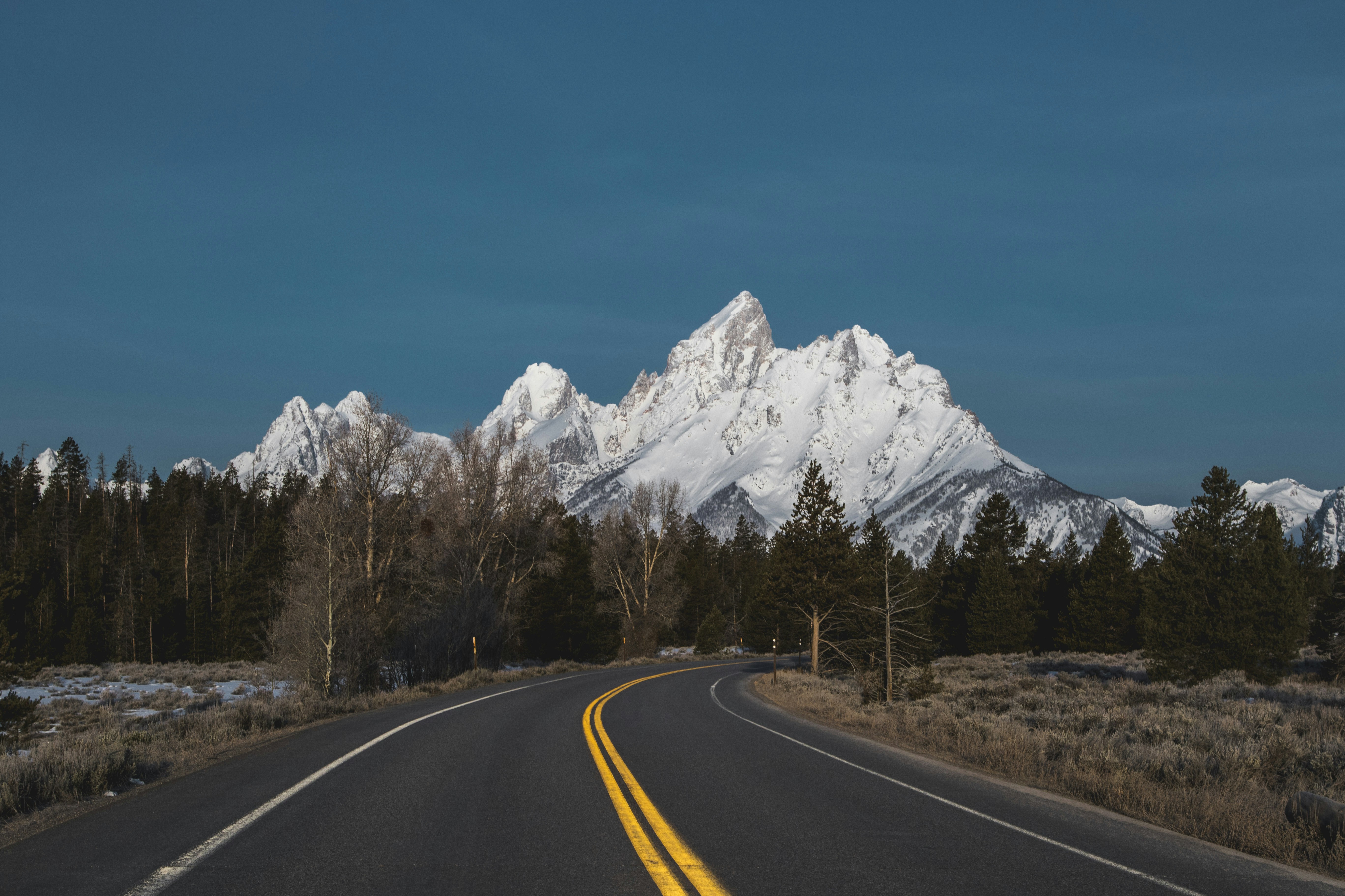 Snow-capped Grand Teton mountains rise prominently behind a winding road under a clear blue sky.